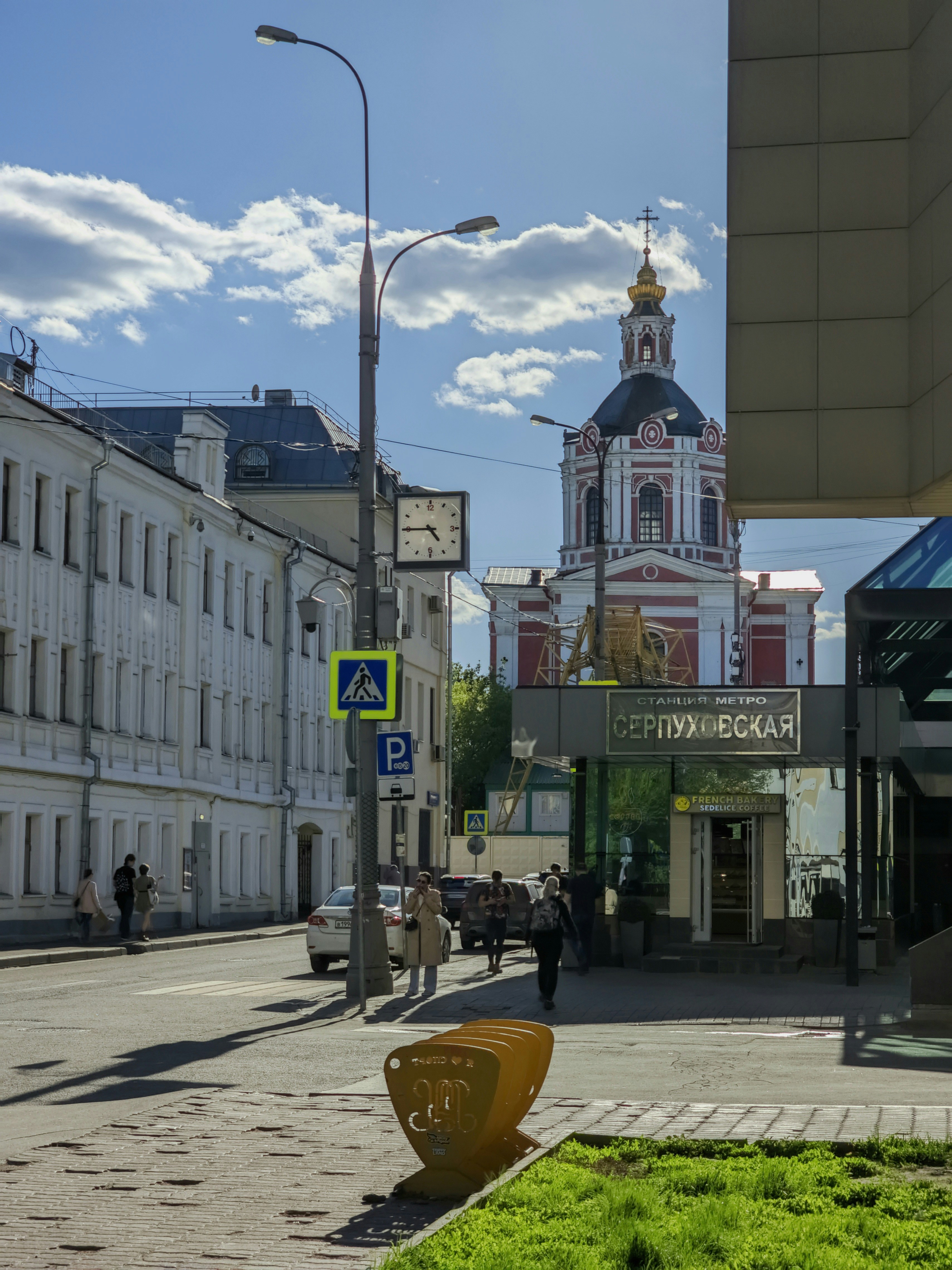 Sunlit Moscow street scene with a red-and-white church capped by a blue dome in the background and a nearby metro entrance.