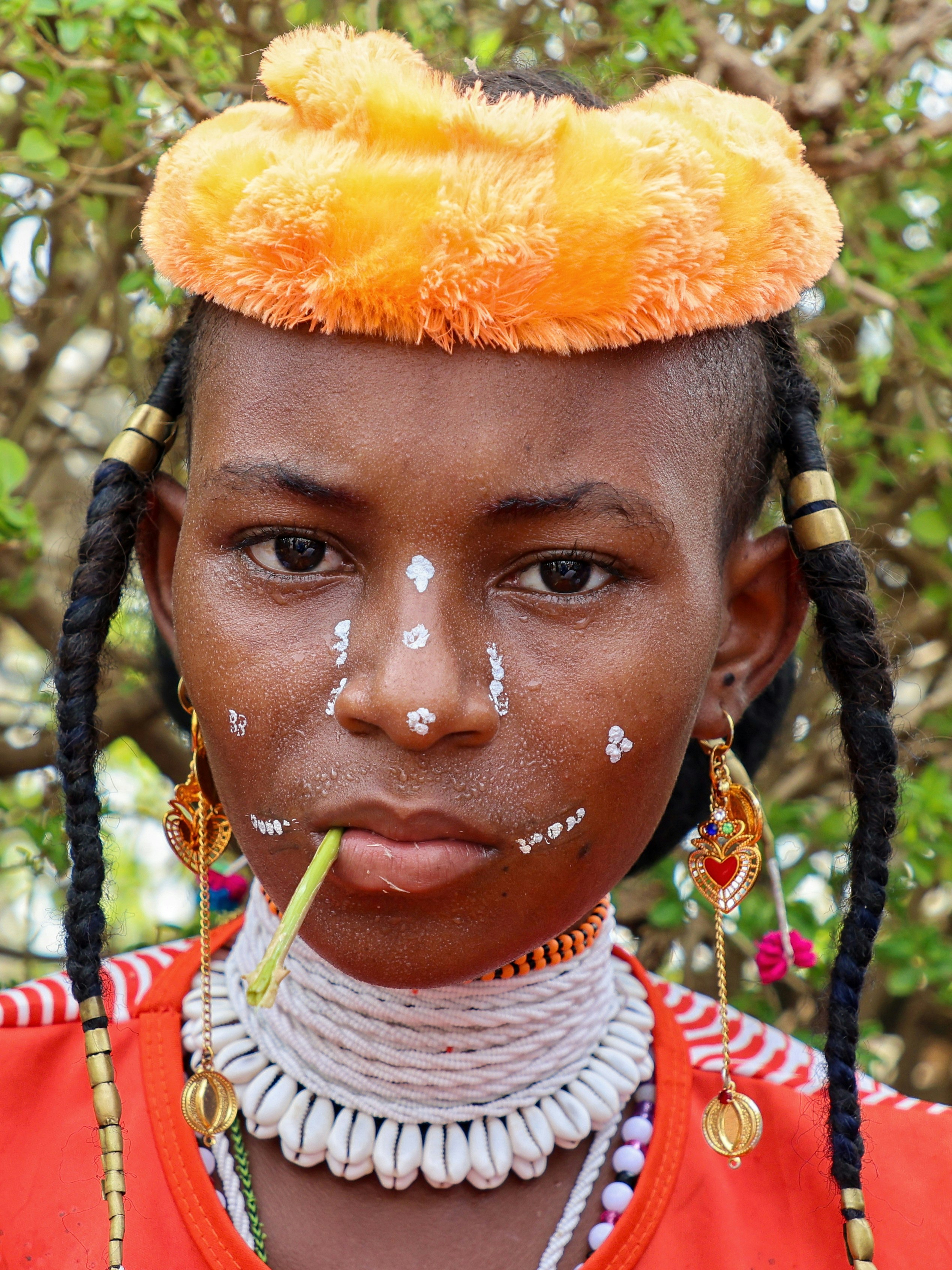 Close-up photograph of a young wearer adorned with white face paint, shell jewelry, and a vivid orange headpiece, set against lush greenery.