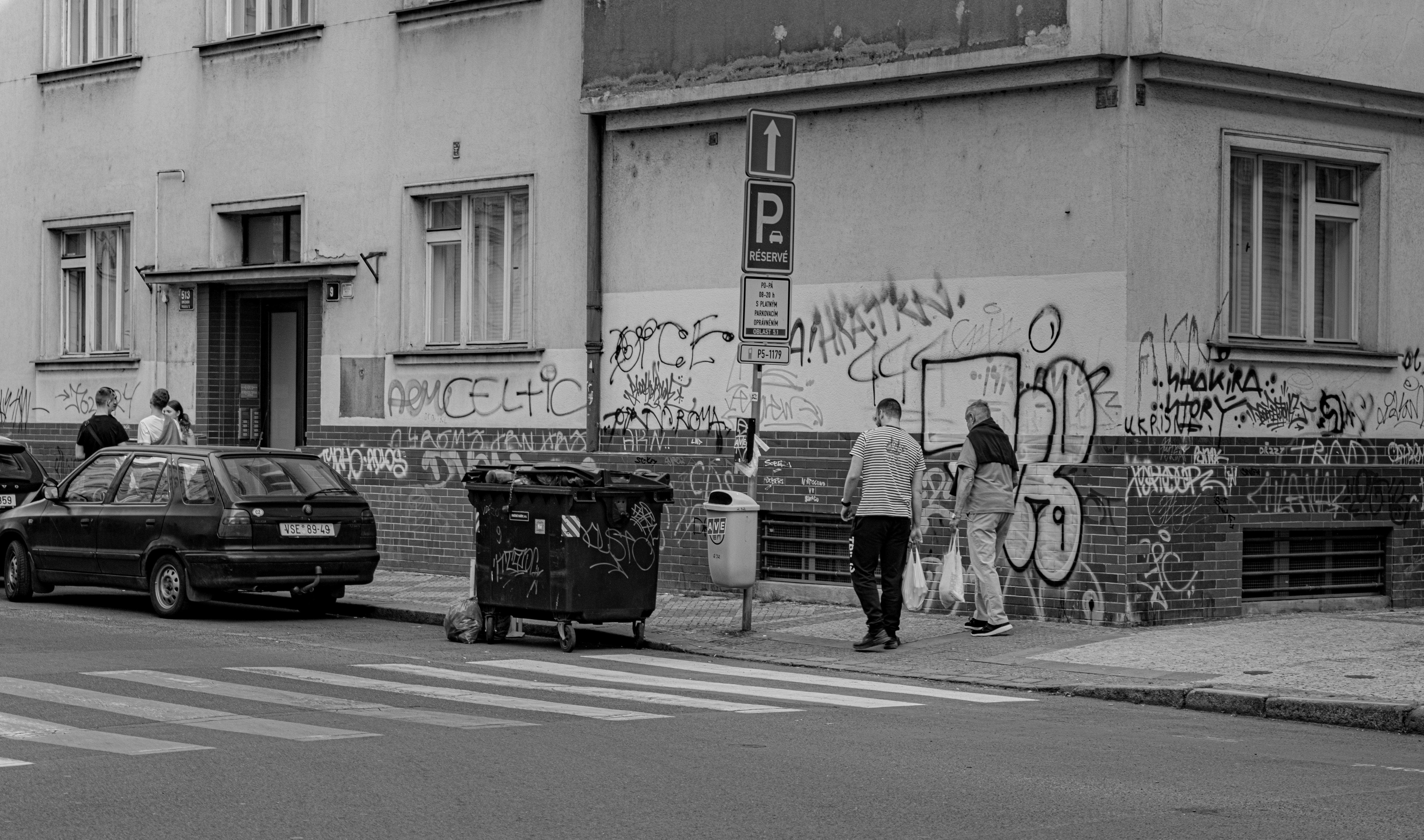 Graffiti-covered building corner with pedestrians walking by and a parked car in a city environment.