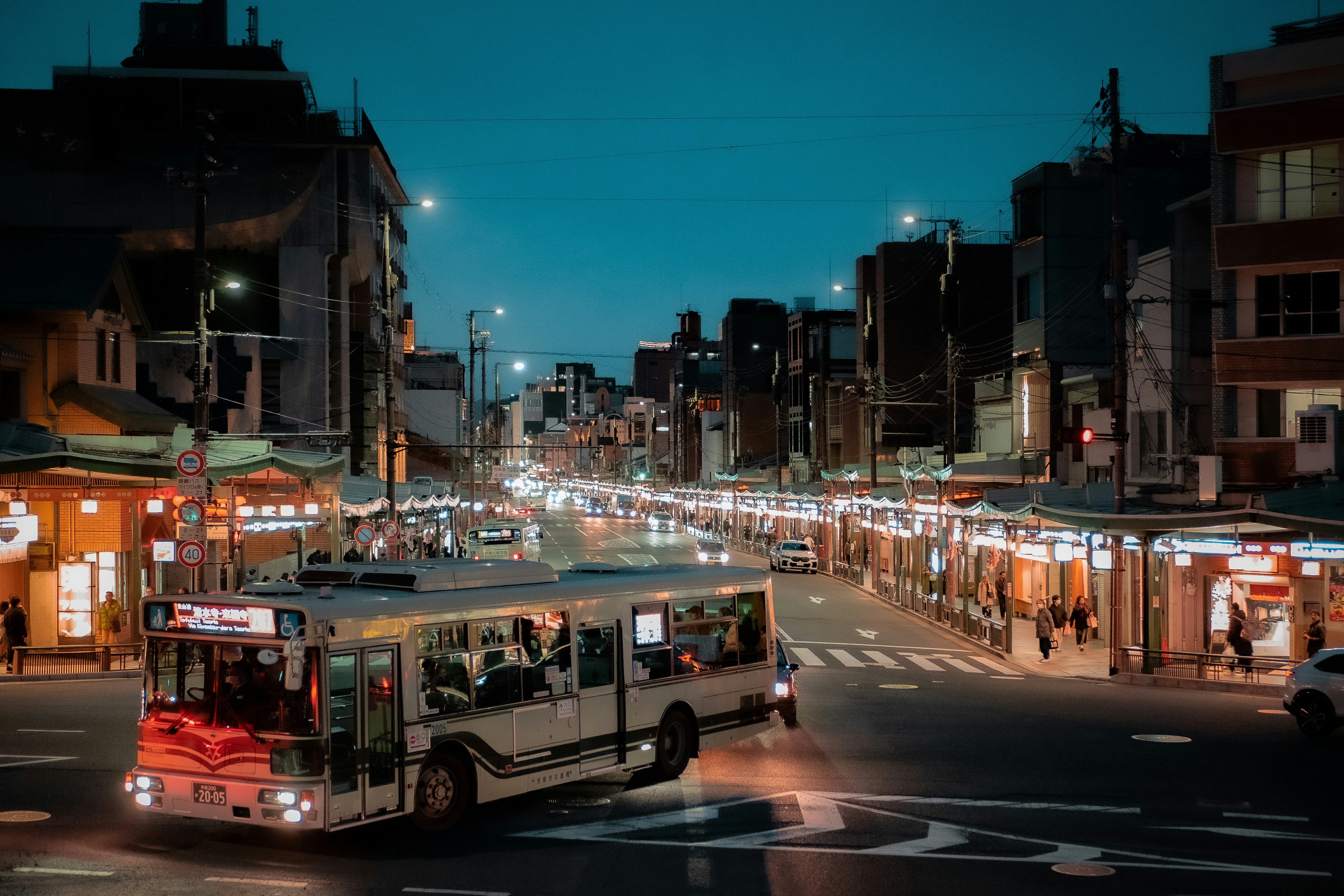 City bus navigating a busy intersection as twilight descends, with illuminated storefronts lining the street.