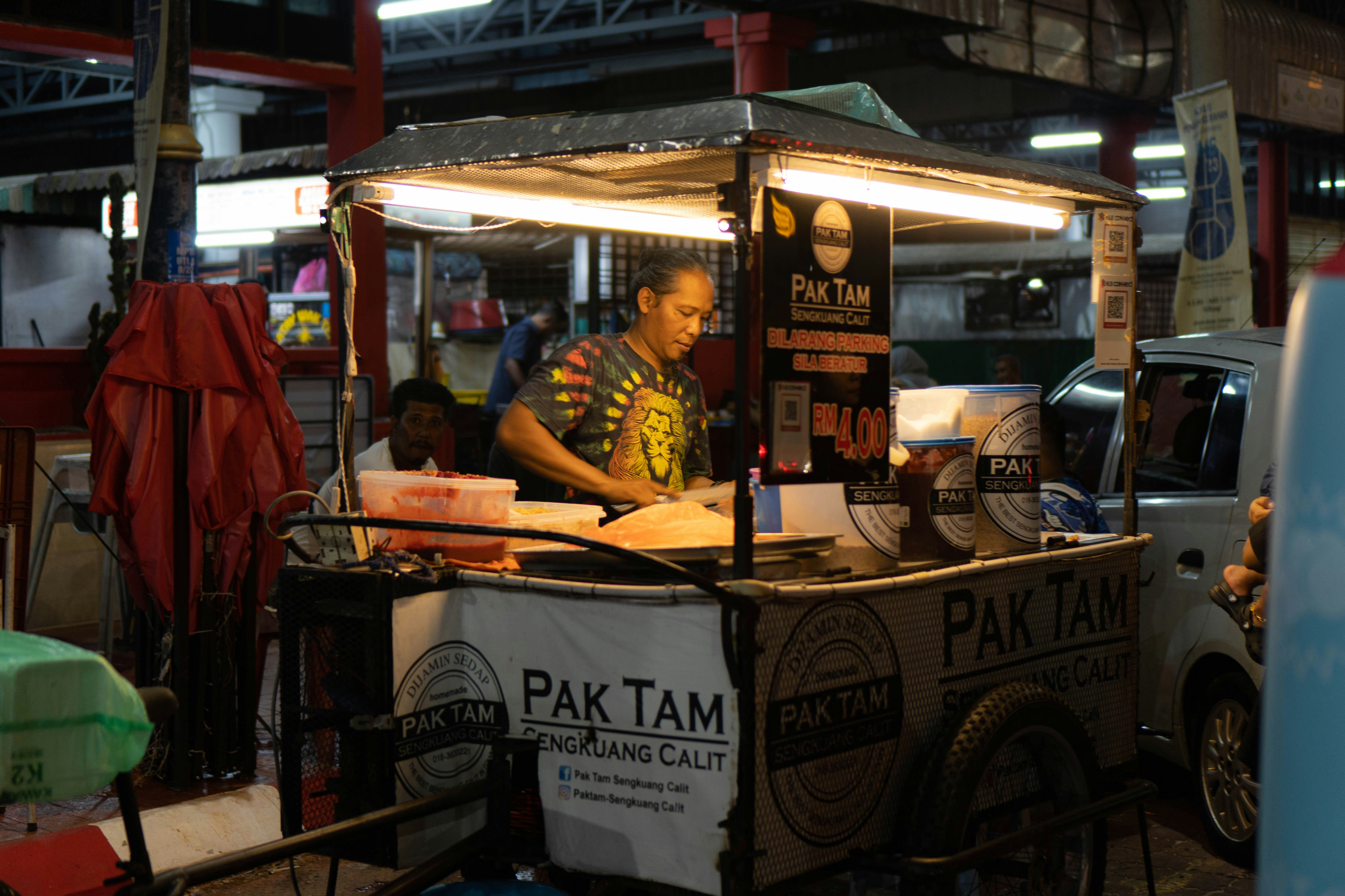 Vendor preparing food at a brightly lit street food cart in a bustling night market.