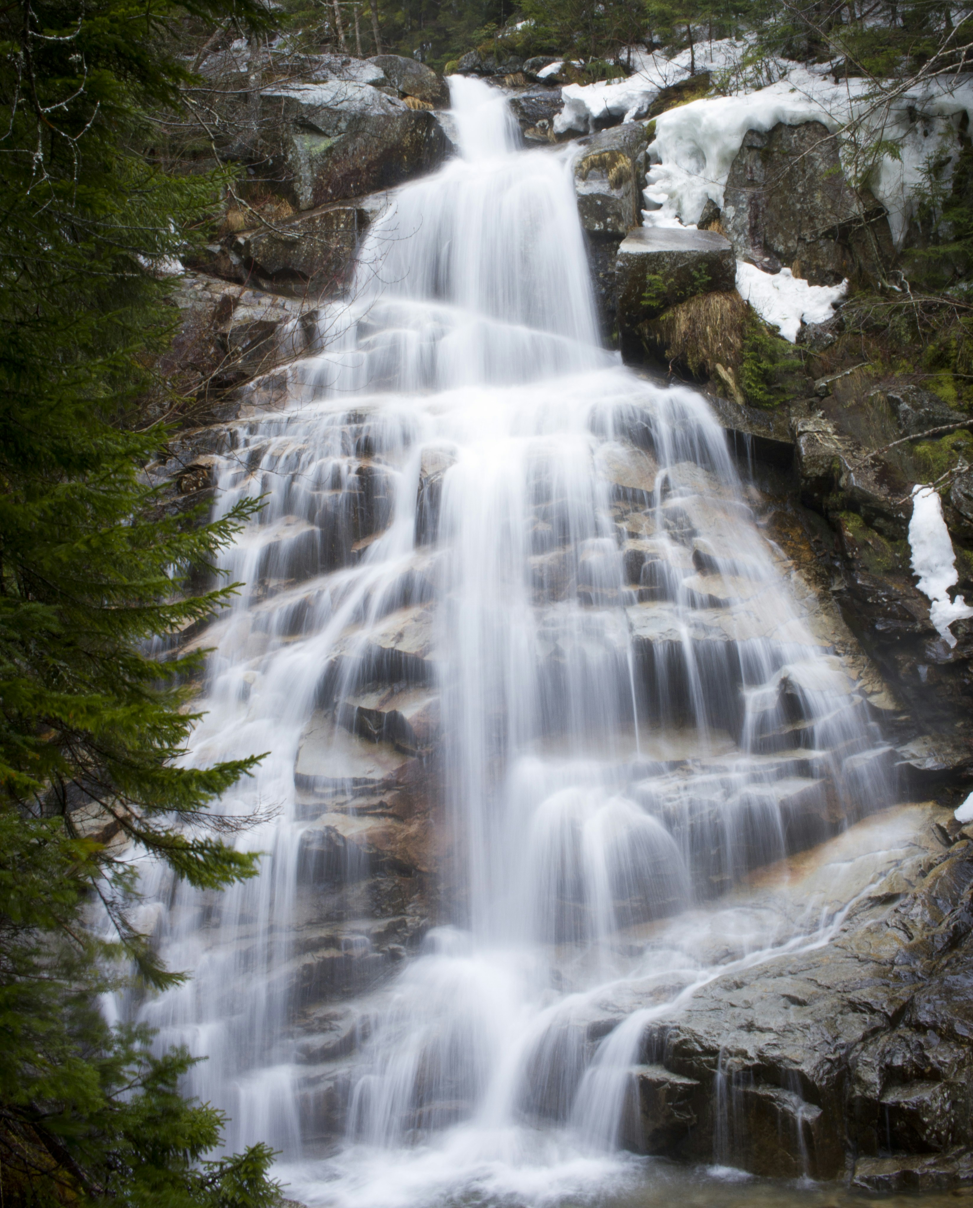 Gentle waterfall cascading over rocky terrain, surrounded by lush greenery and remnants of snow. The scene captures the tranquility of nature's flow.
