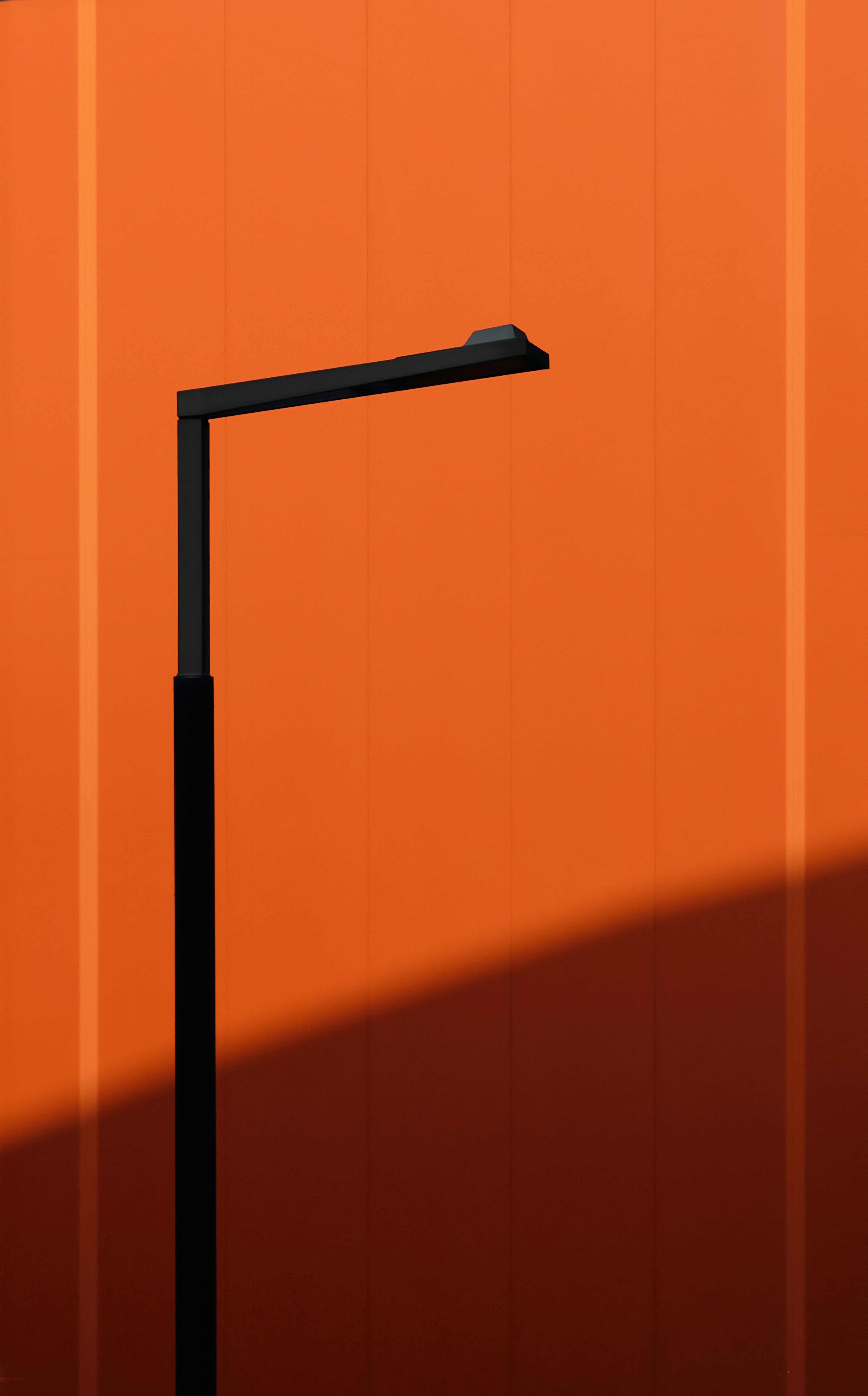 A solitary streetlamp rises against a vivid orange wall, its clean silhouette interrupted by a diagonal shadow across the lower panel.