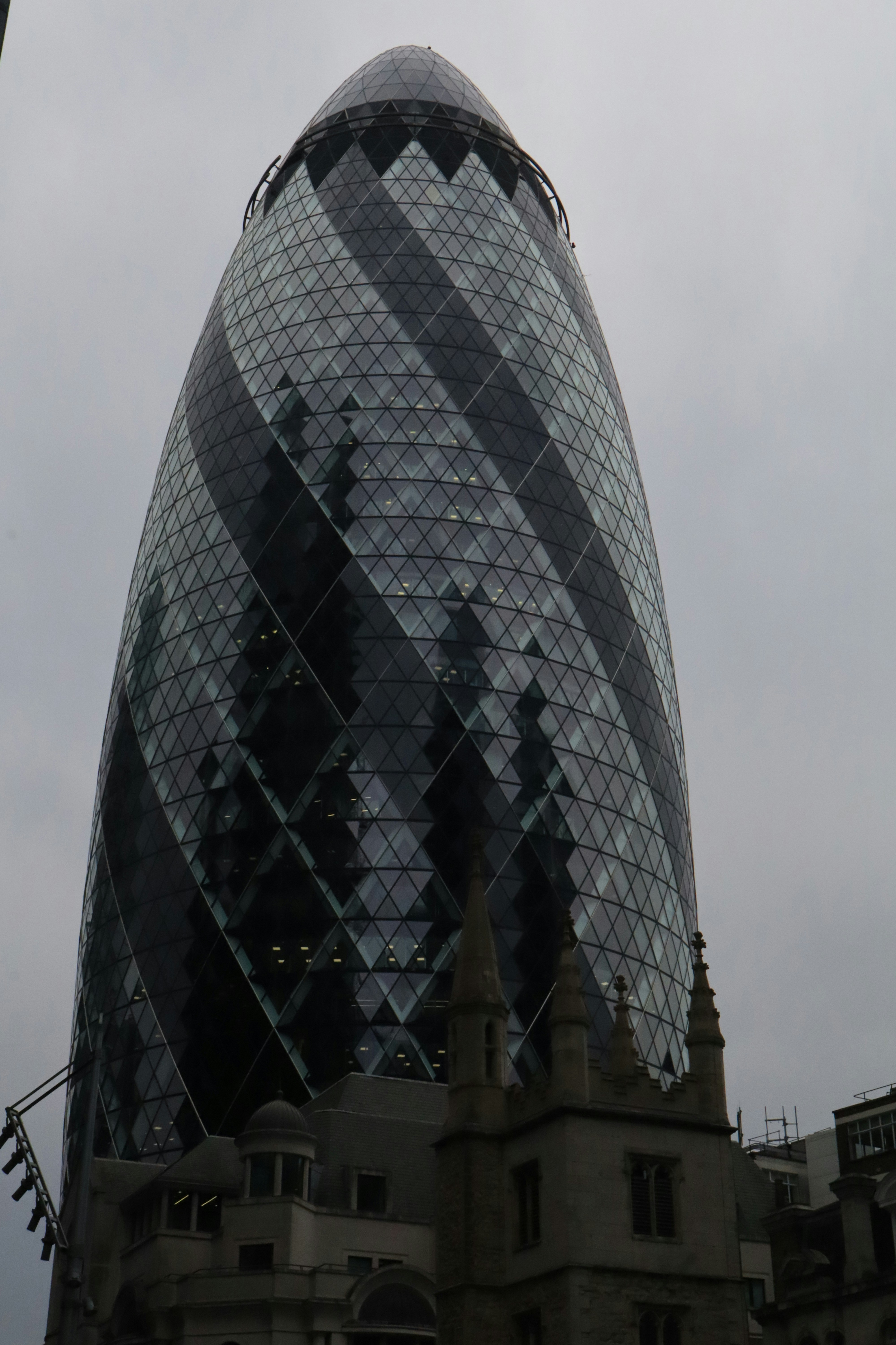 Photograph of The Gherkin (30 St Mary Axe) skyscraper towering over historic rooftops under a gray, overcast sky. The glass-diamond facade contrasts with the older architecture at street level.