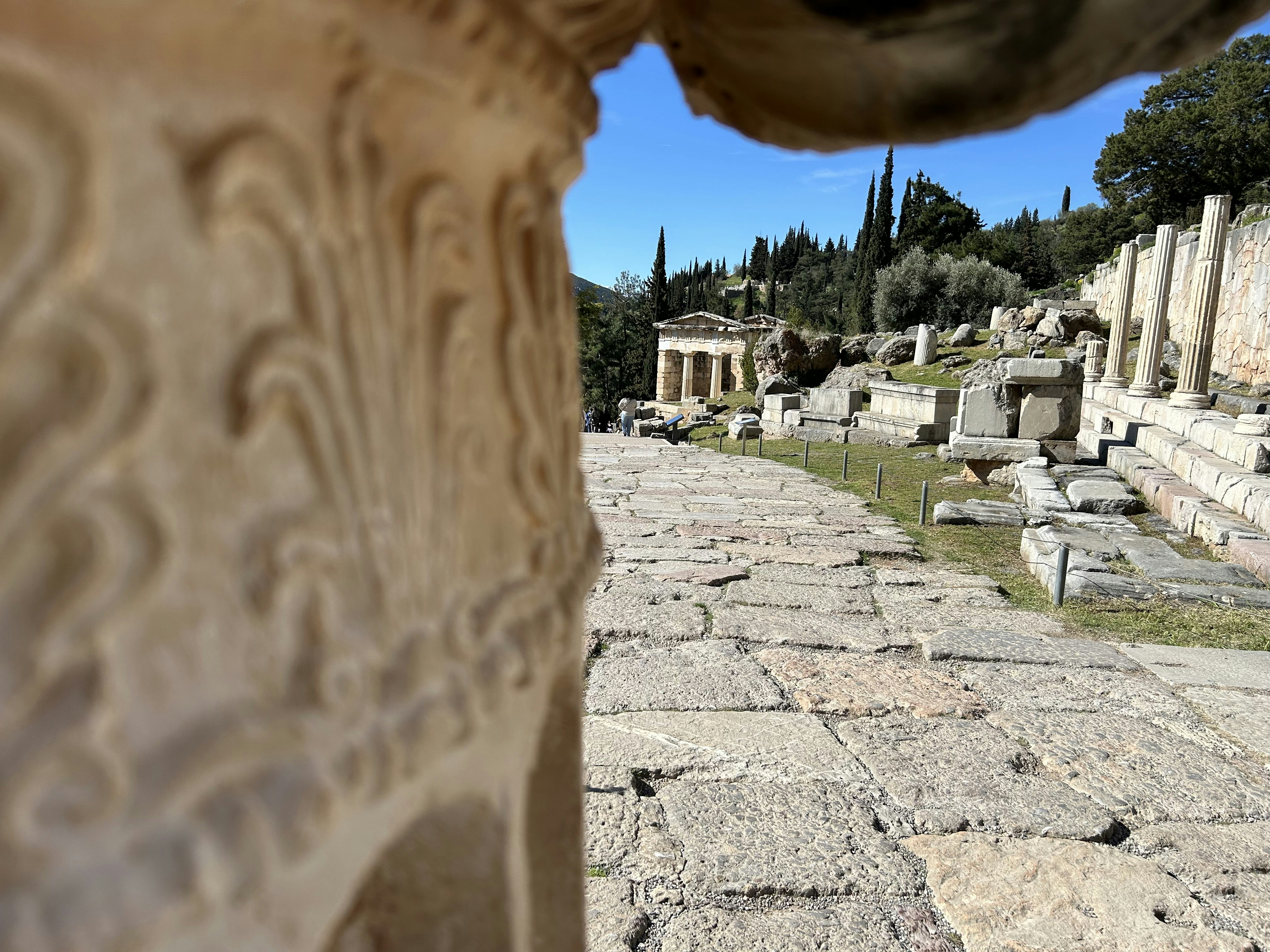 Ancient stone pathway leading to a historical structure, framed by intricately carved stonework in the foreground. Sunlight illuminates the ruins, highlighting their weathered beauty.