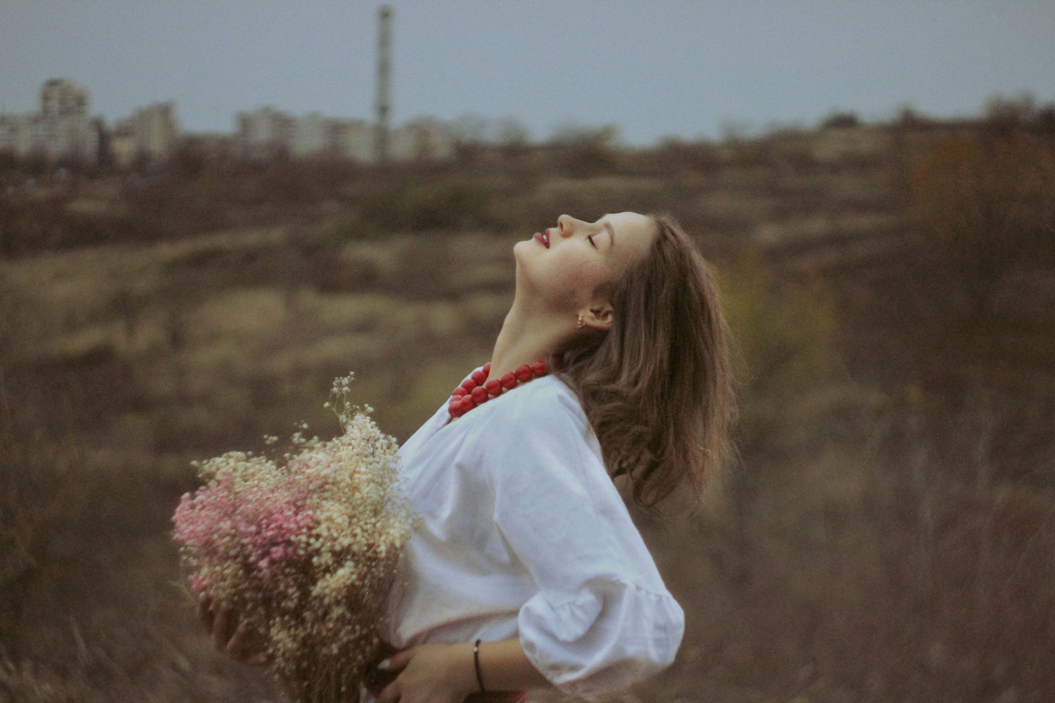Woman in white blouse holding flowers, standing in a field with her head tilted back.