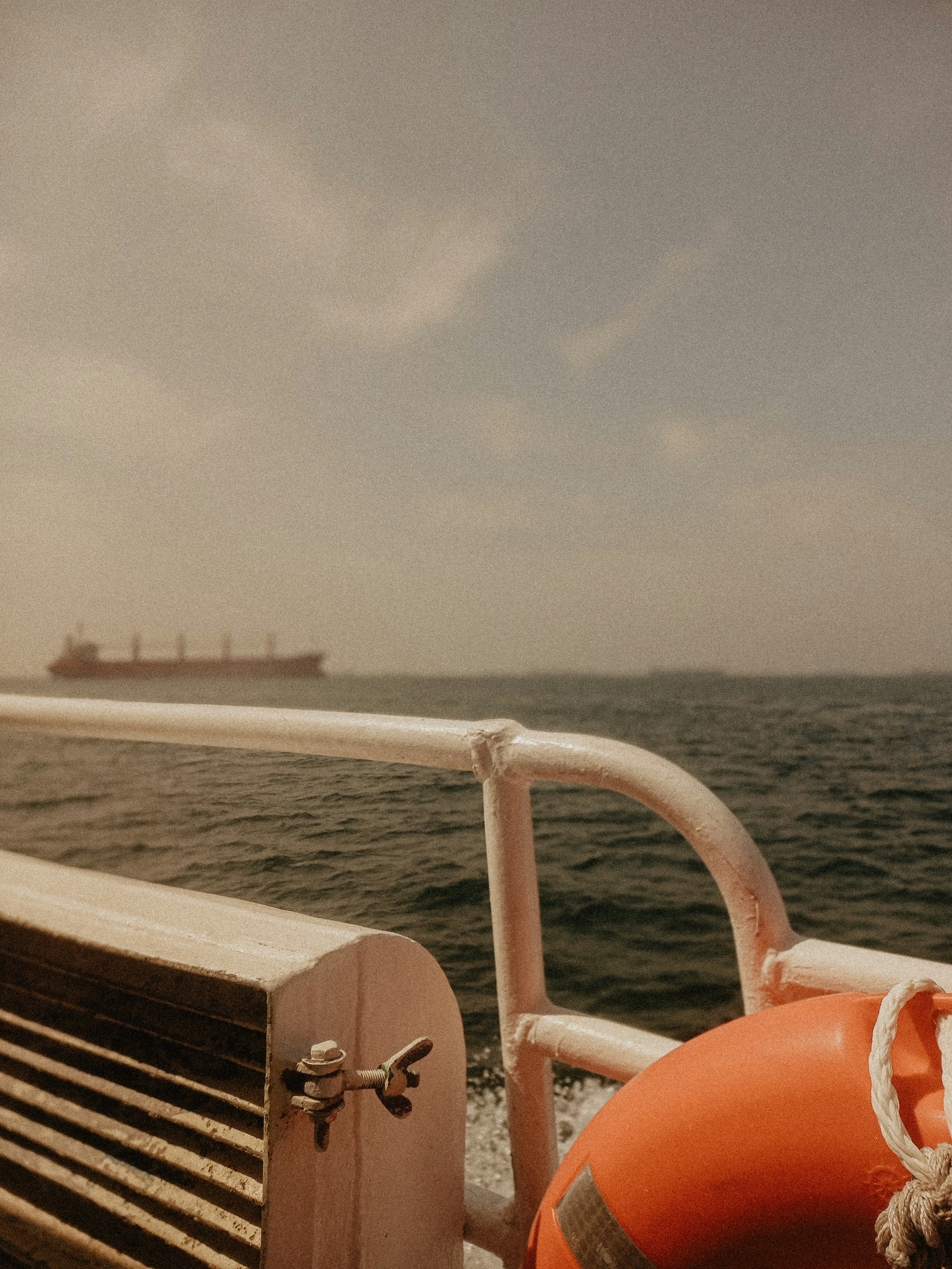 Orange life ring attached to a ship railing frames the foreground as the calm sea leads to a distant freighter on the horizon.