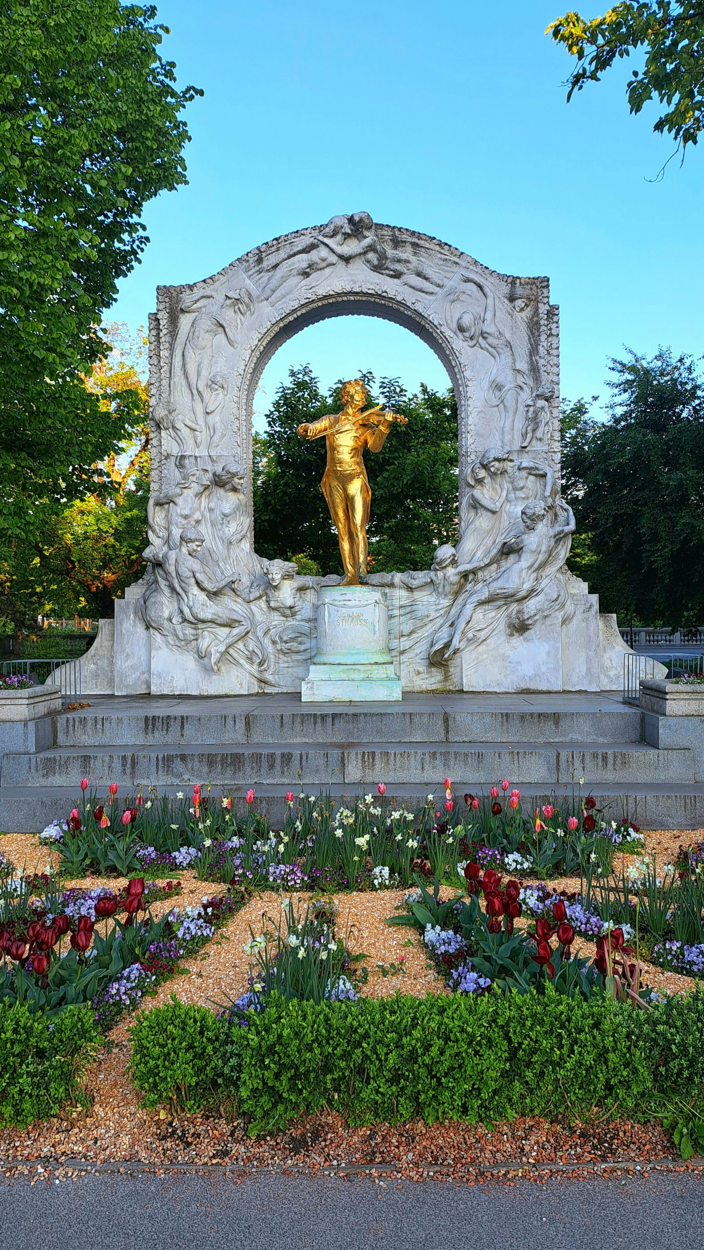 Golden statue of a violinist framed by a sculpted arch, surrounded by vibrant tulip gardens.