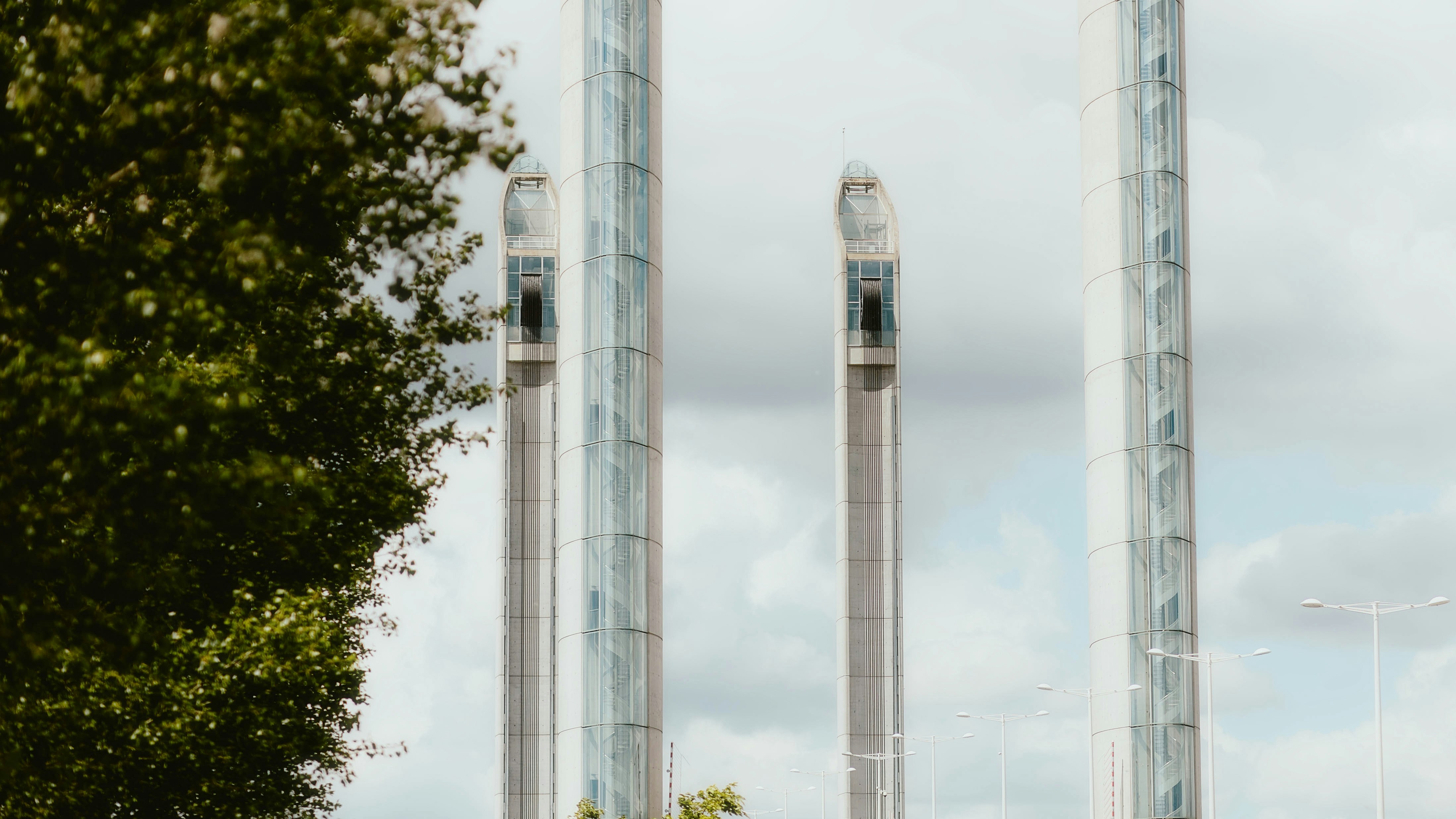 Tall glass towers rise against a backdrop of cloudy sky, flanked by lush green trees.