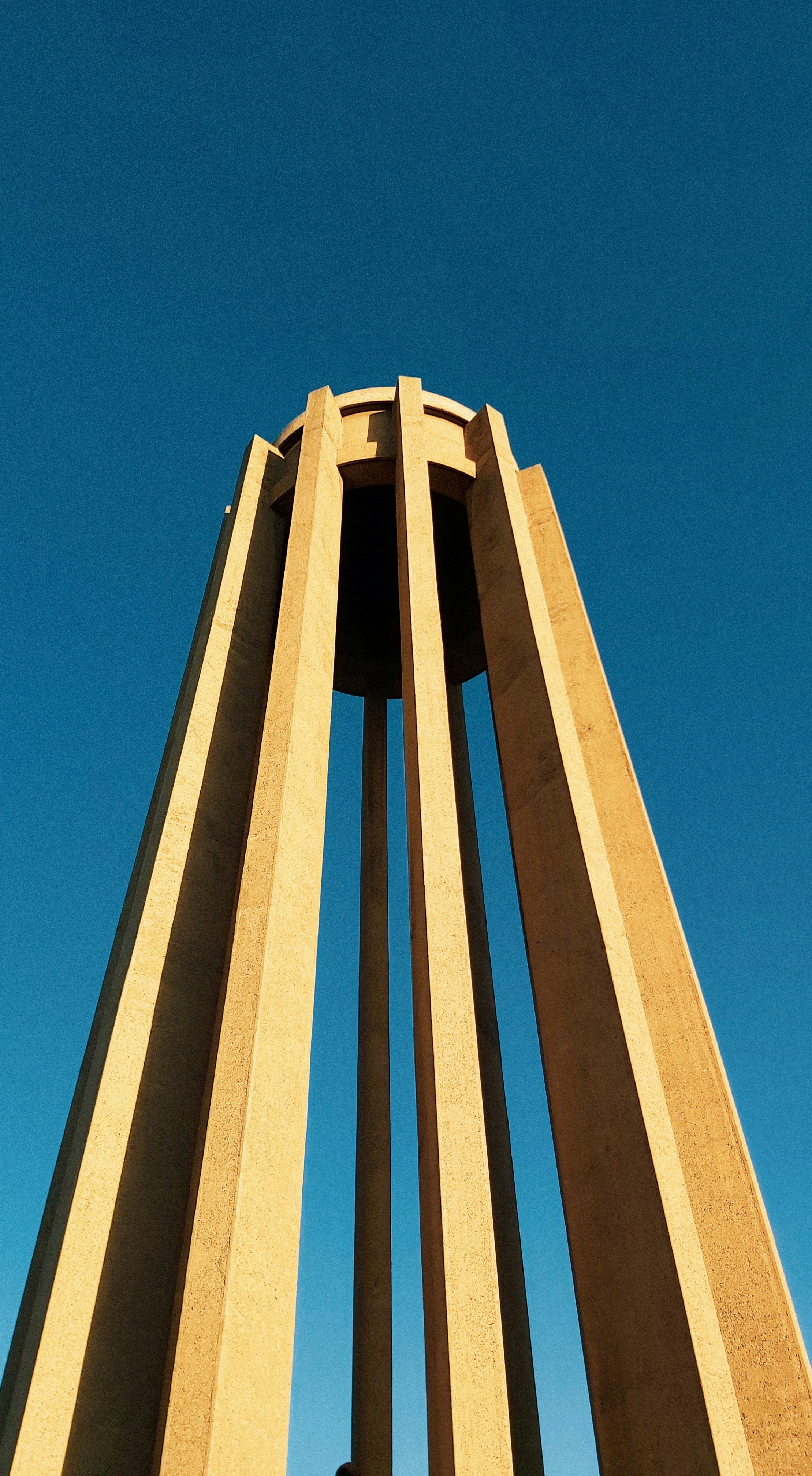 Tall concrete monument with elongated vertical columns forming a circular opening at the summit, set against a deep blue sky.