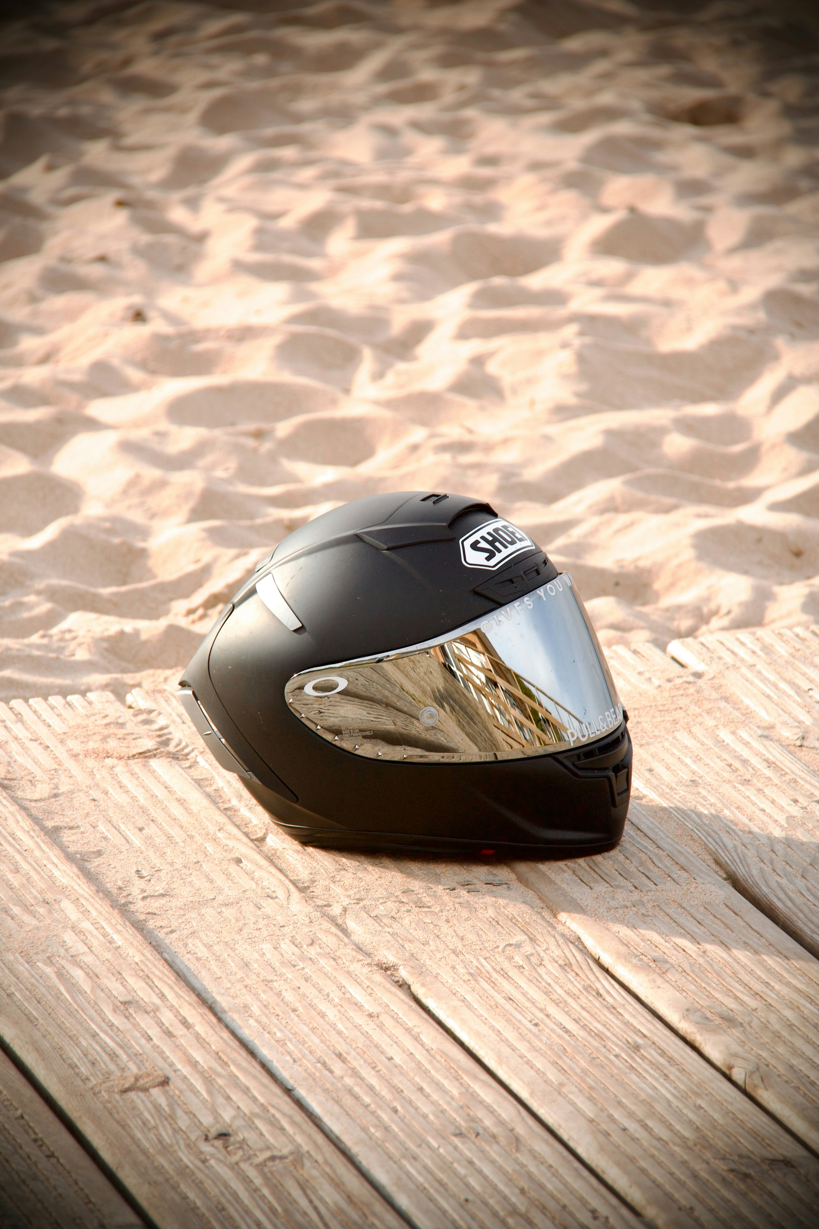 A solitary SHOEI helmet rests on sunlit wooden planks at the edge of the sandy boardwalk