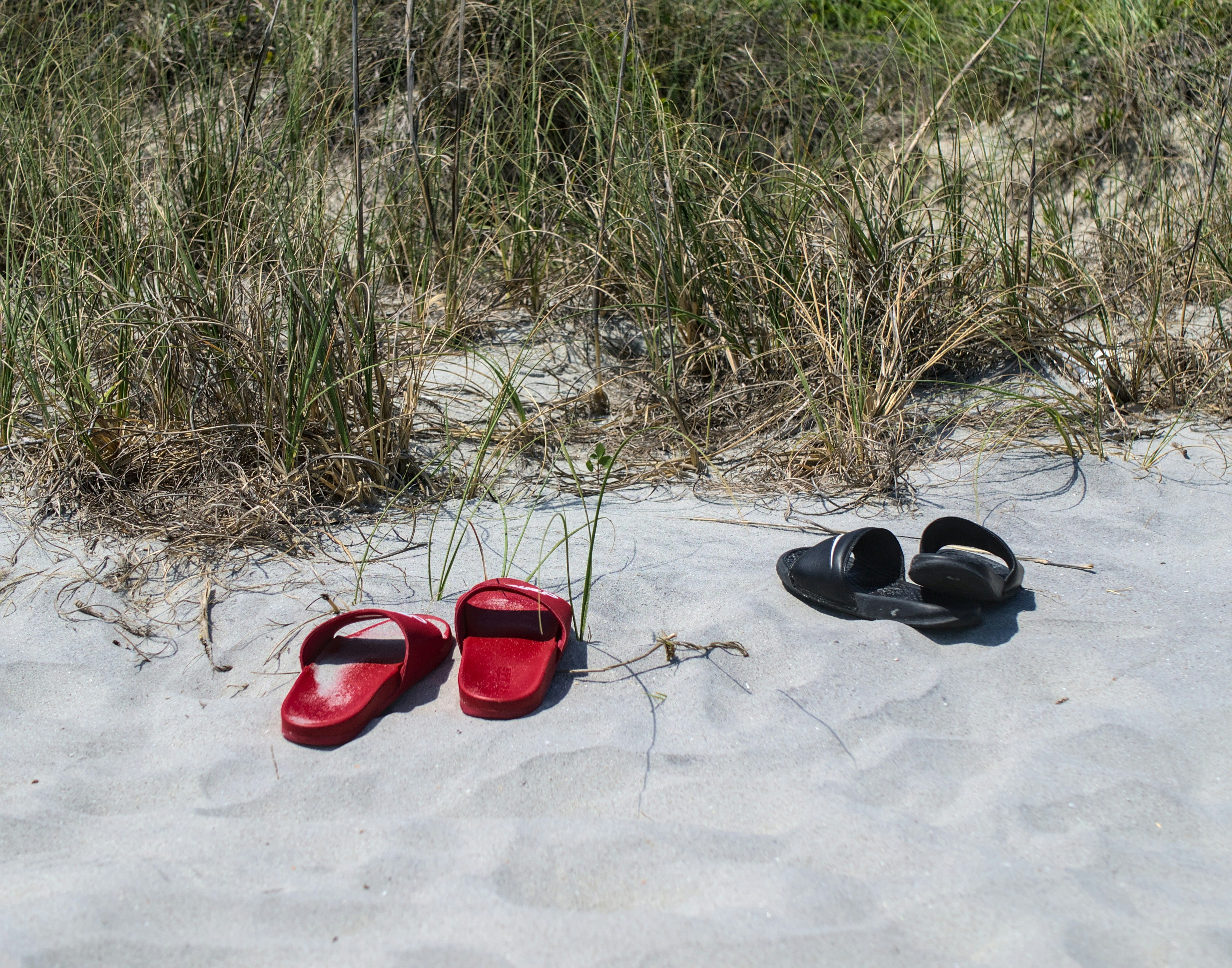 Two pairs of sandals, red on the left and black on the right, rest on sunlit white sand with dune grass behind in a simple beach photograph.