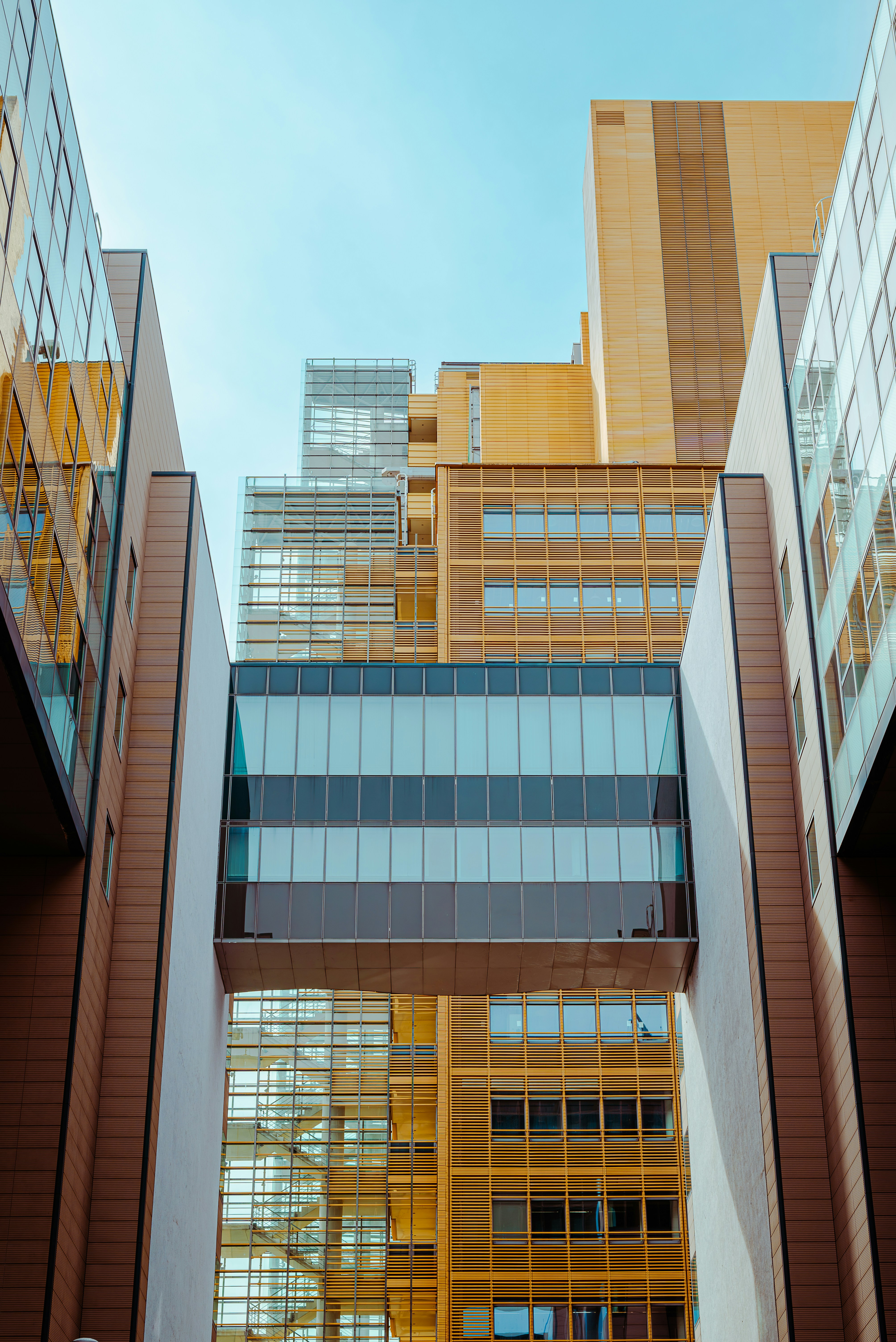 Architectural photograph of a glass skybridge nestled between golden-tinted towers against a bright blue sky. The composition emphasizes reflective surfaces and geometric lines.