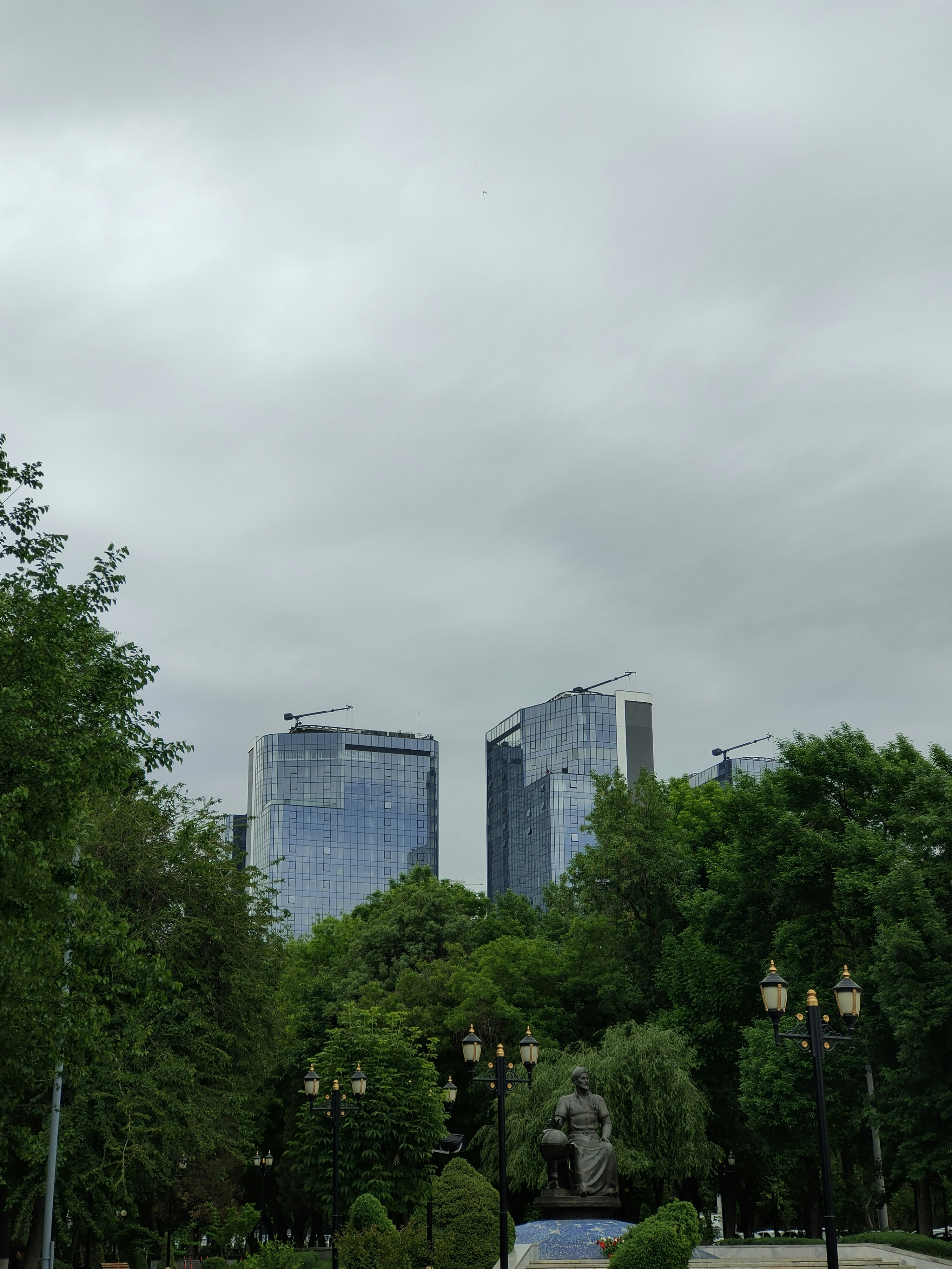 A stone statue sits amid trees and vintage lampposts in a city park, with modern glass skyscrapers rising behind.