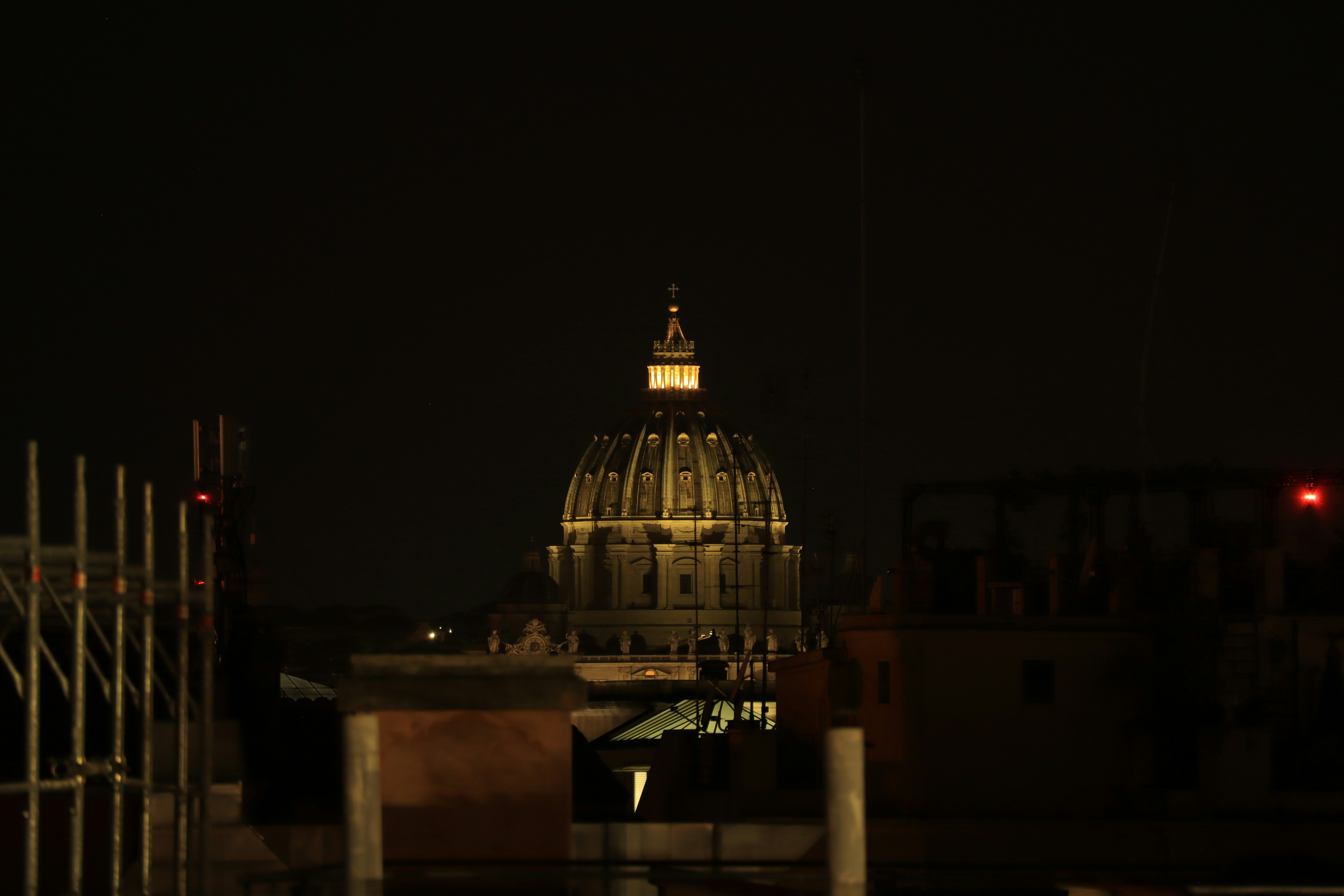 The dome of St. Peter's Basilica illuminated against the night sky, framed by rooftops and structures of Rome.