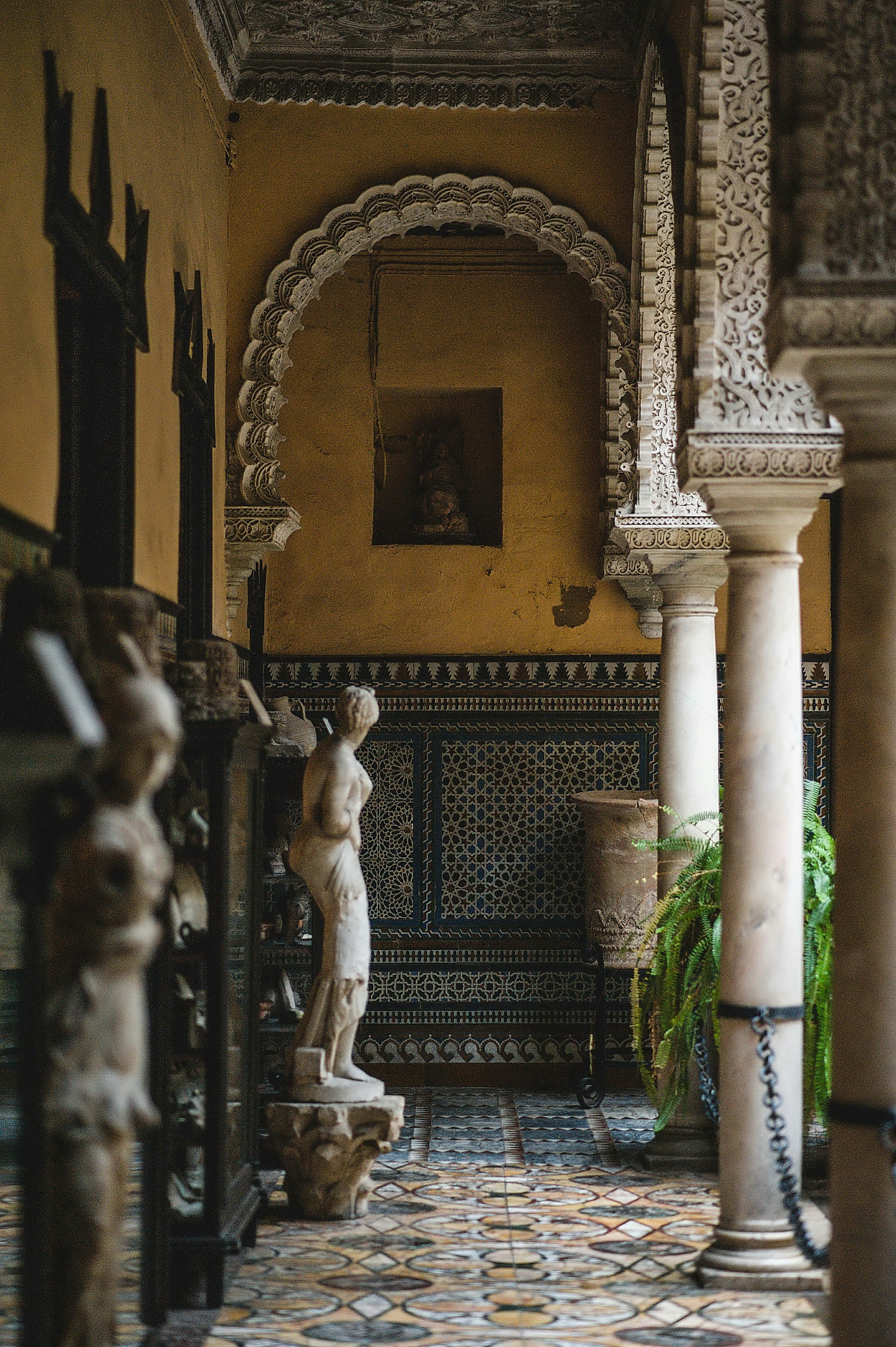 A classical statue stands in a sunlit Moorish courtyard, flanked by ornate arches and carved columns. The scene captures a quiet interior passage with patterned tile flooring and potted plants.