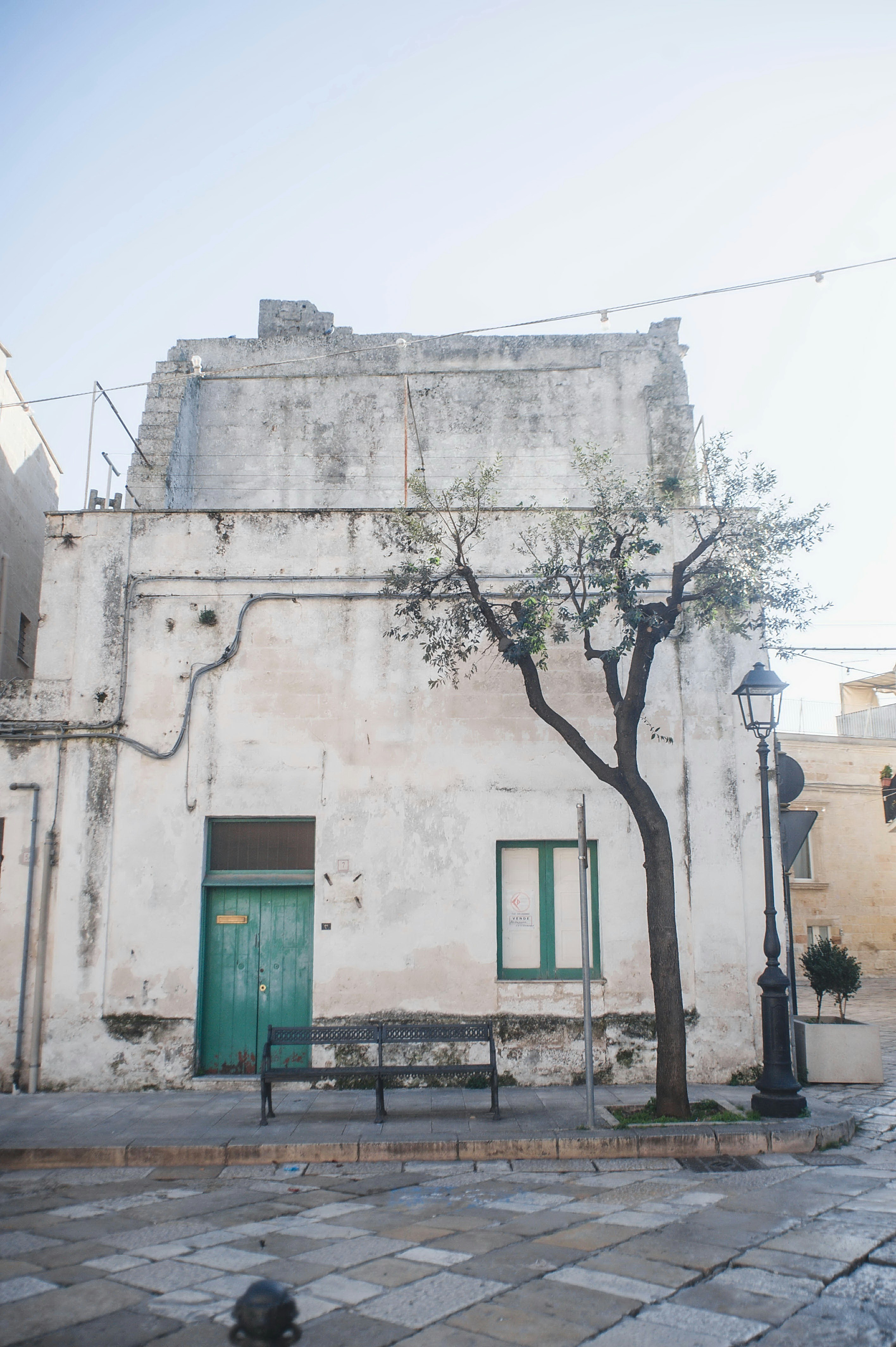 Weathered building with a green door and window, adjacent to a lone tree and bench in a sunlit plaza.