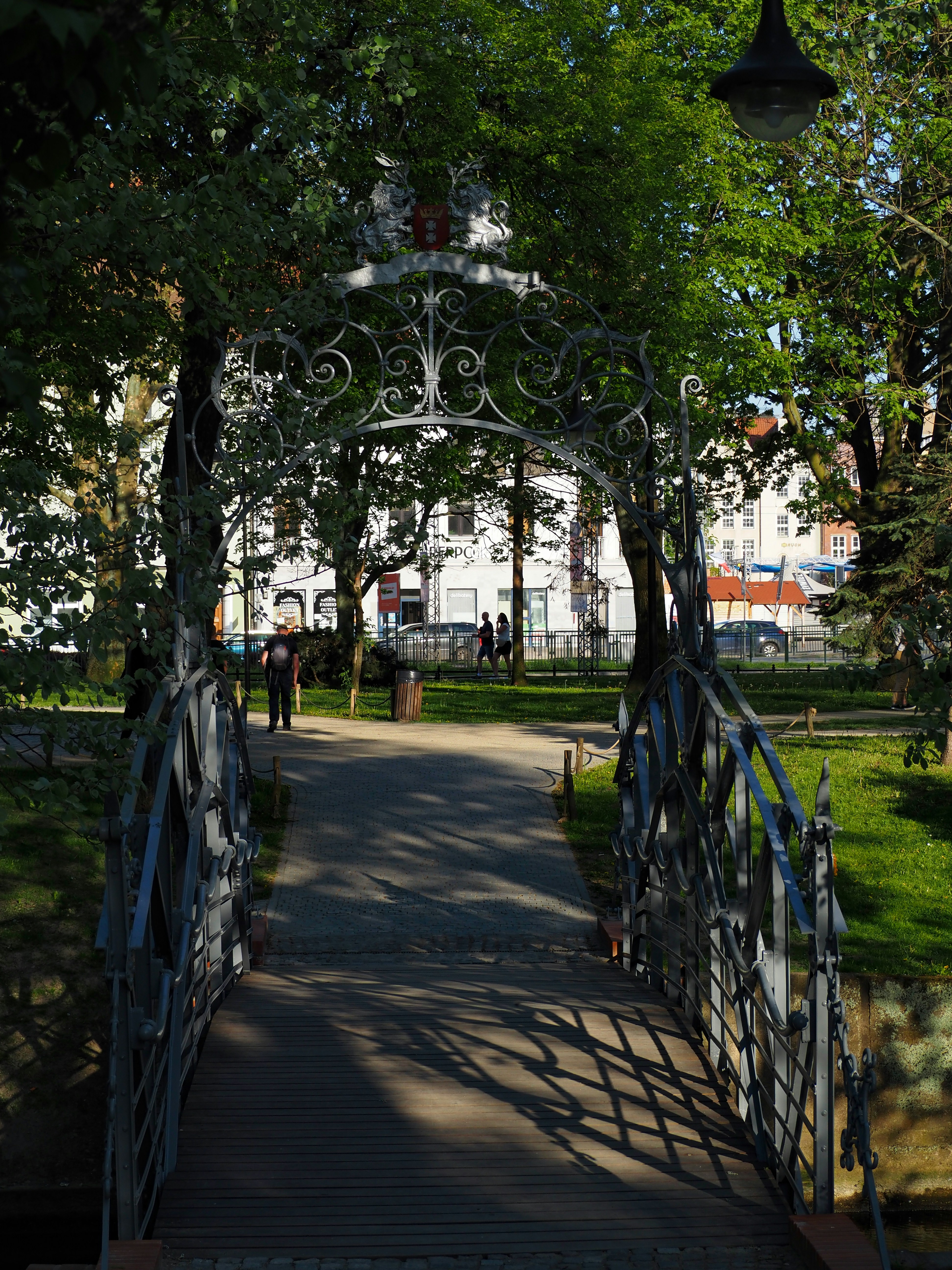 Wrought iron bridge arching over a serene path, surrounded by lush greenery and dappled sunlight. Pedestrians stroll in the background, adding life to the tranquil scene.