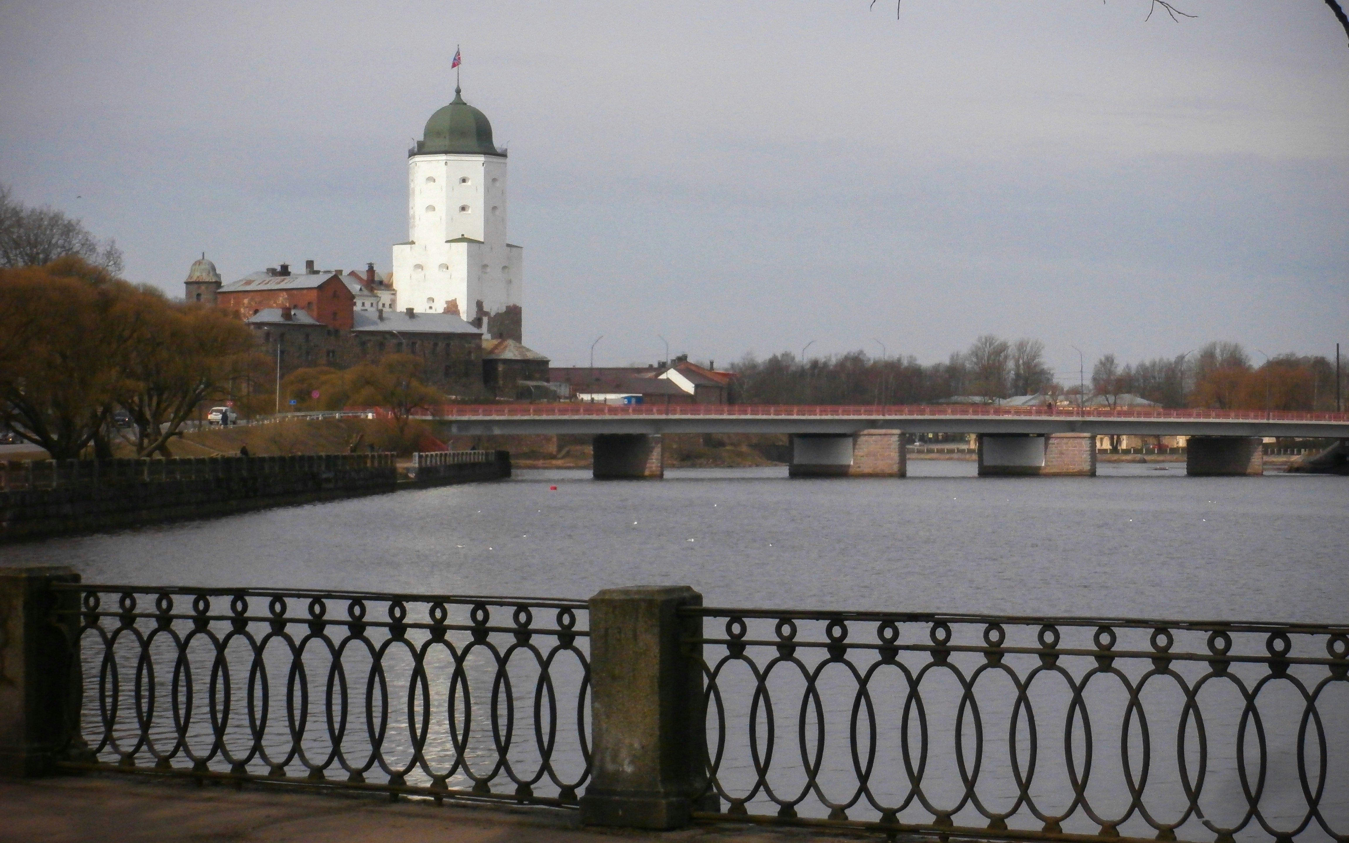 Riverside view of an old fortress with a prominent white tower, framed by a decorative railing and a distant bridge.