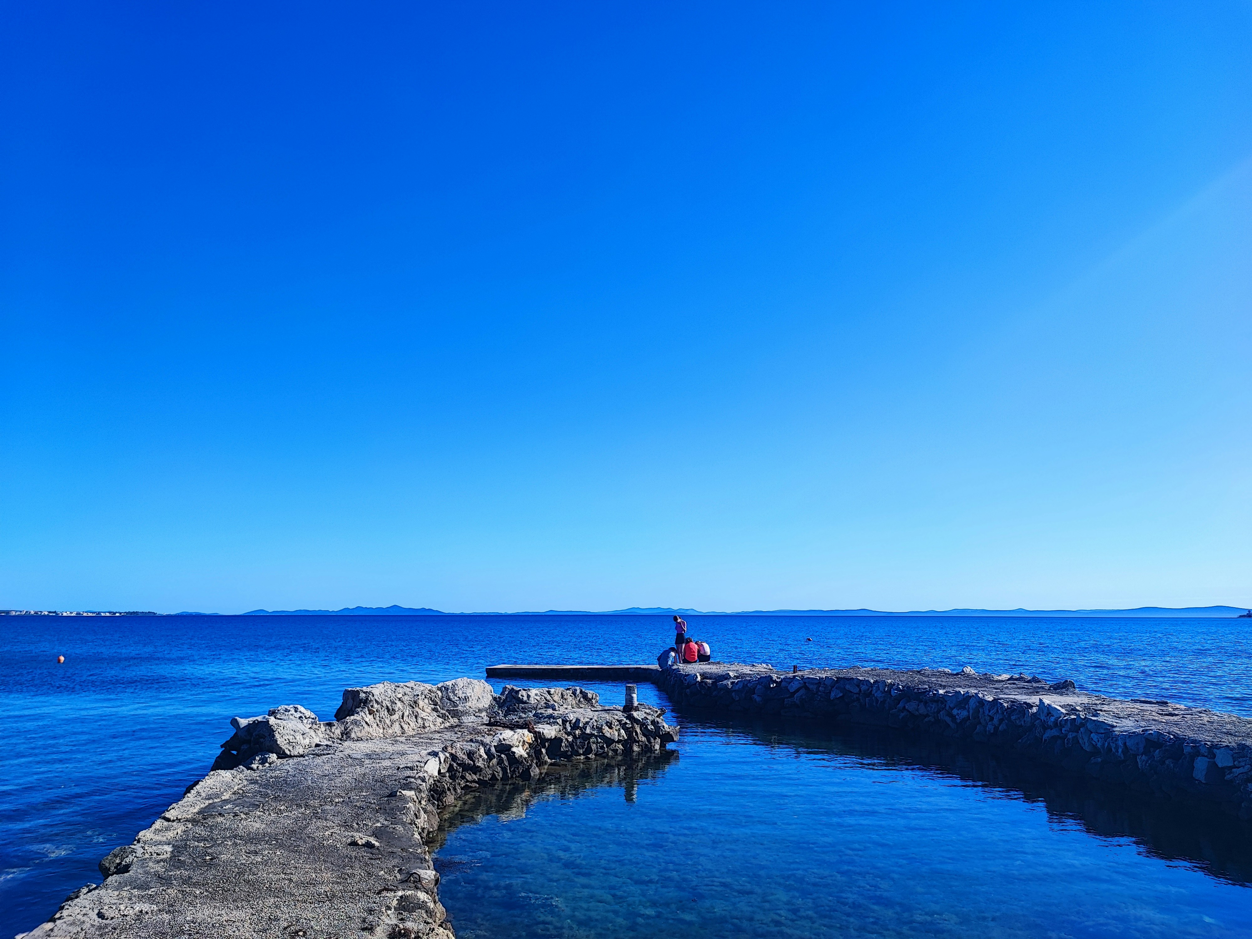 Calm blue sea stretches to the horizon as a curved stone jetty leads to a lone figure at its tip. The bright, cloudless sky completes the tranquil coastal scene.