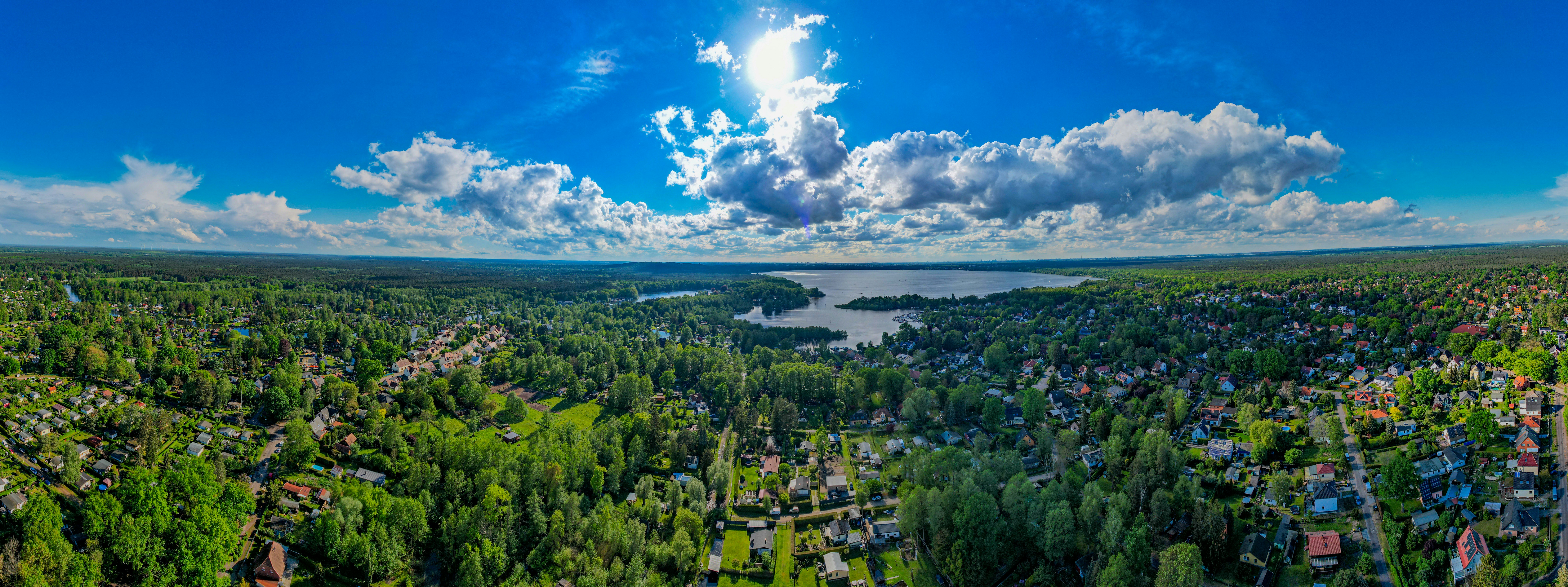 This panoramic aerial view captures the Müggelsee area in Berlin, showcasing a mix of residential neighborhoods and lush greenery around a large lake. The image depicts dense tree cover interspersed with houses and roads, under a clear sky with prominent clouds. It highlights the scenic, tranquil vibe of the suburb, blending natural and urban elements seamlessly in a well-developed community.