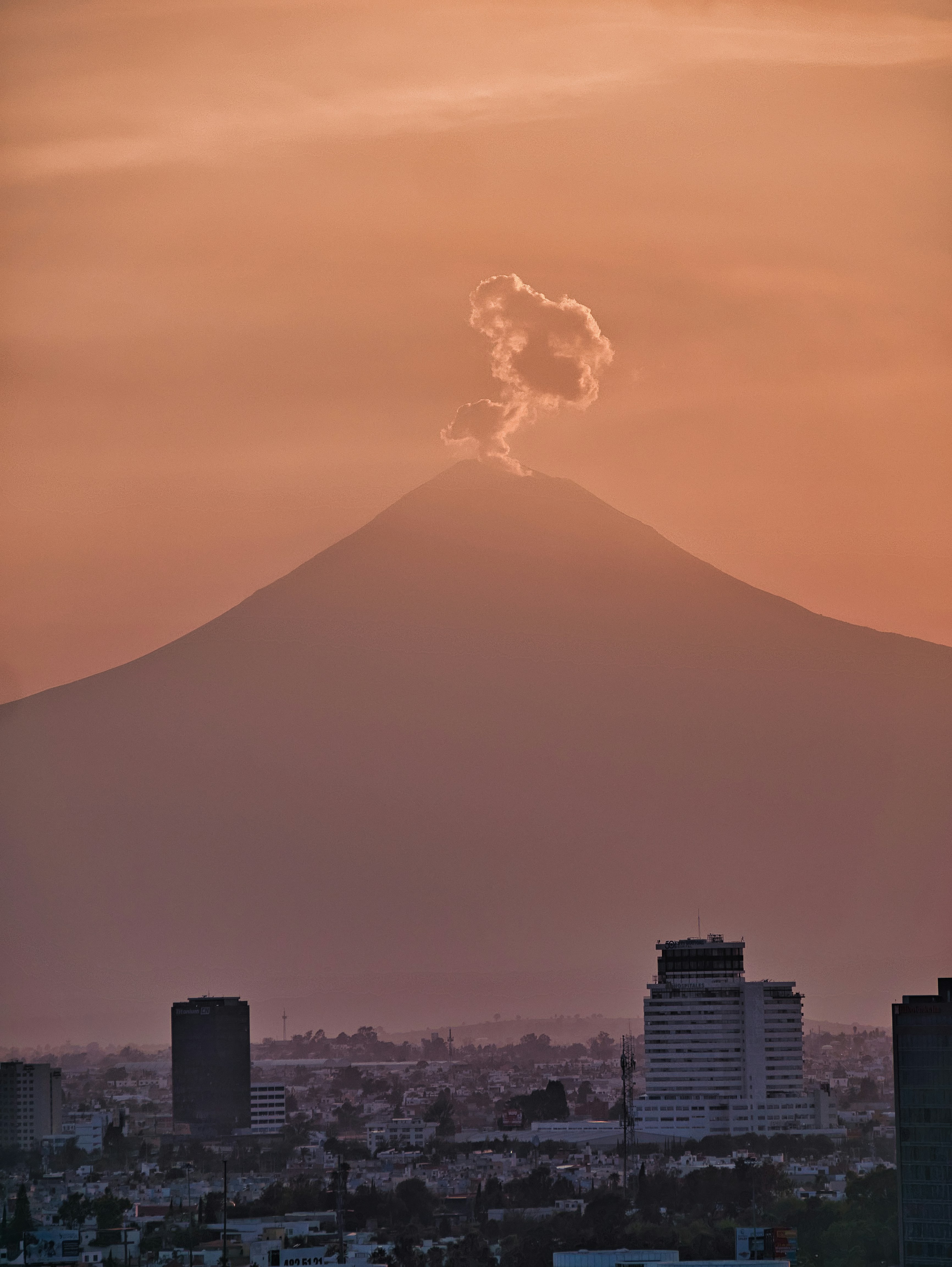 Sunset photograph of a distant volcano with a plume rising above a sprawling city. The warm sky casts silhouettes across the urban foreground, emphasizing scale and distance.