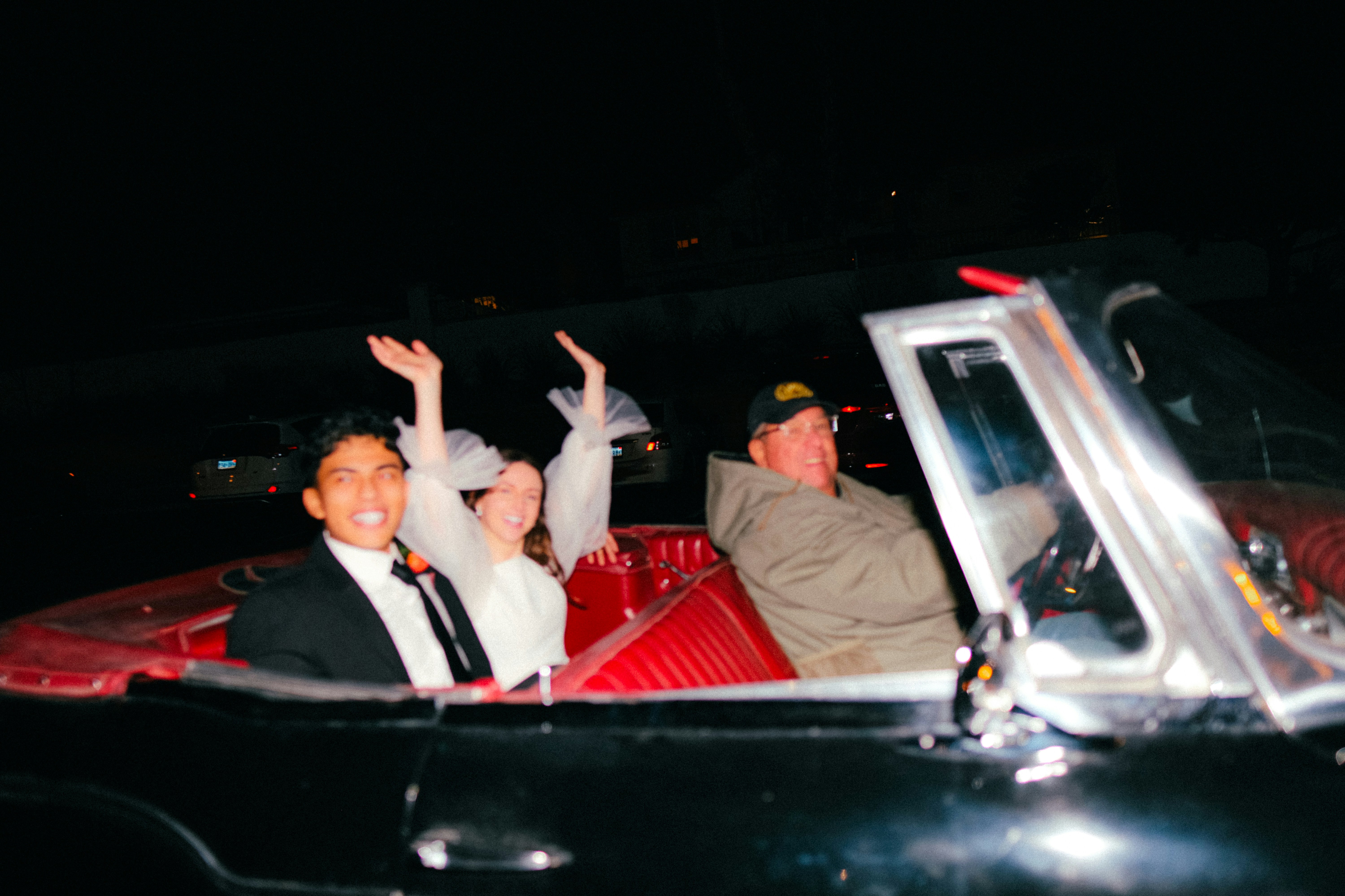 Three people enjoying a ride in a vintage convertible at night, with smiles and raised hands capturing the excitement.
