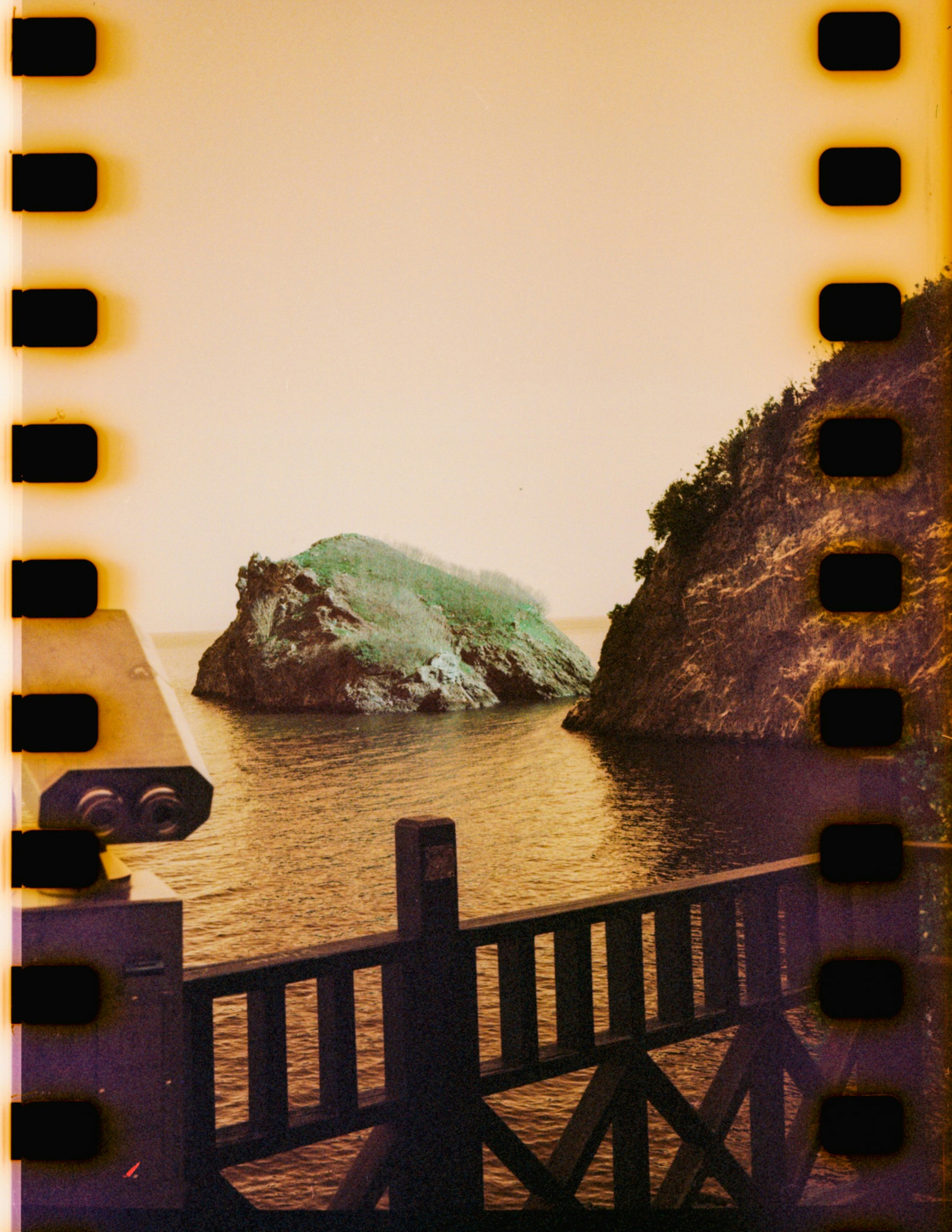 Rock island bathed in warm late-day light sits in calm water, viewed from a wooden railing along a coastal walkway.