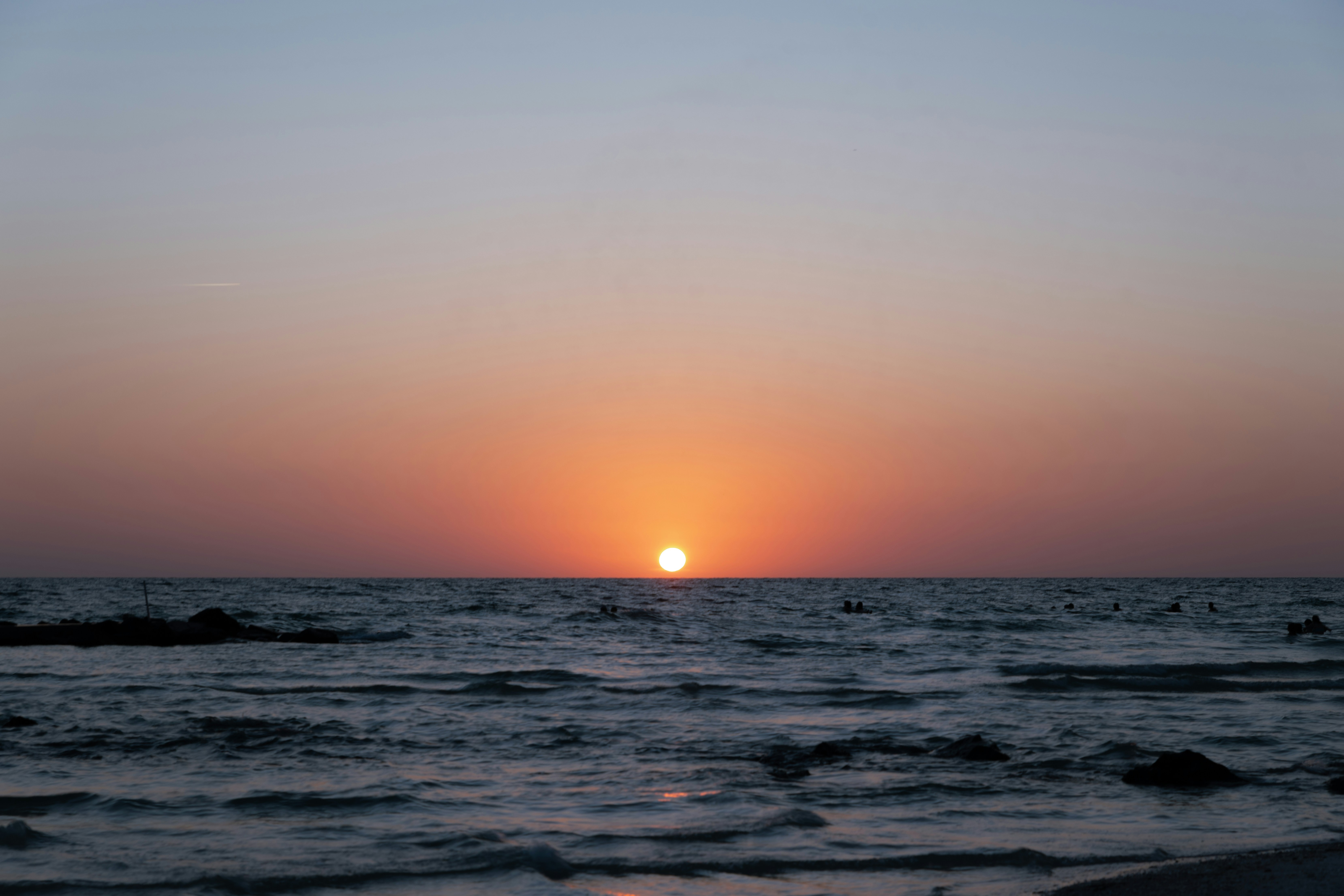 Sun setting over a calm ocean with silhouetted figures in the water.