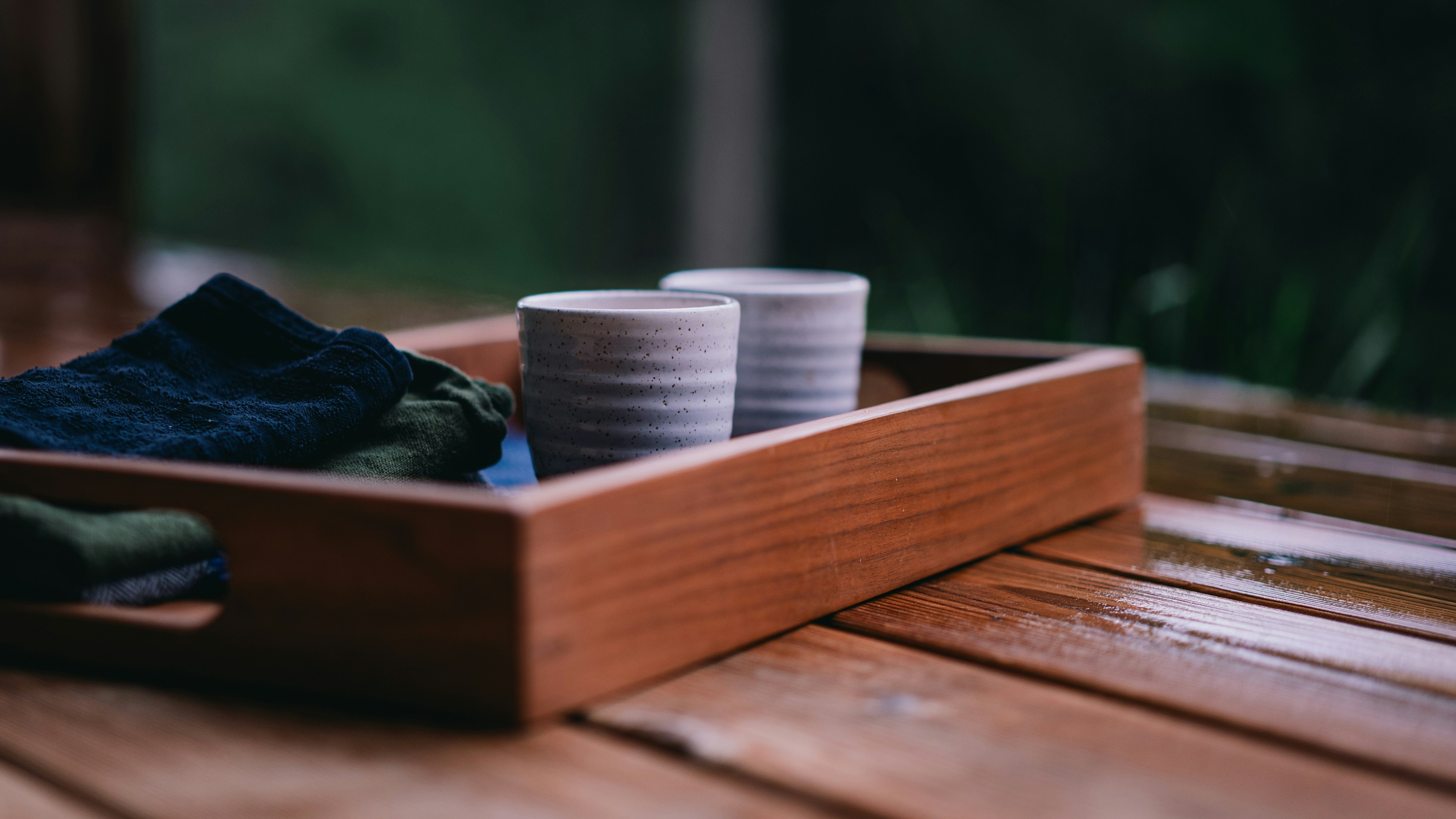 Wooden tray with ceramic cups rests on a rain-wet deck, with folded towels nearby; shallow depth of field softens the background.