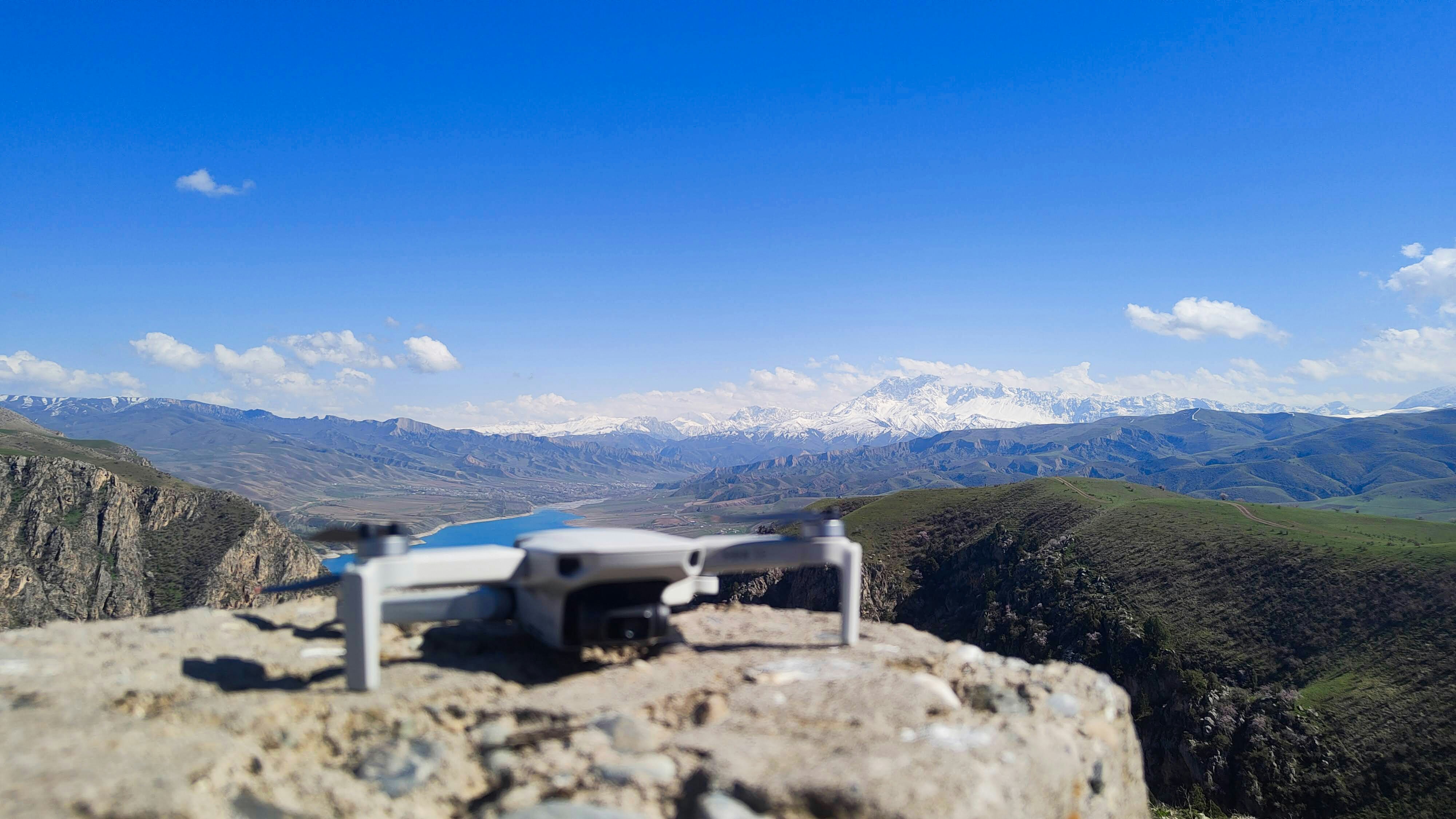 Drone perched on a rugged rock ledge overlooking a vast alpine valley. Snow-capped peaks stretch across the horizon beneath a crisp blue sky.