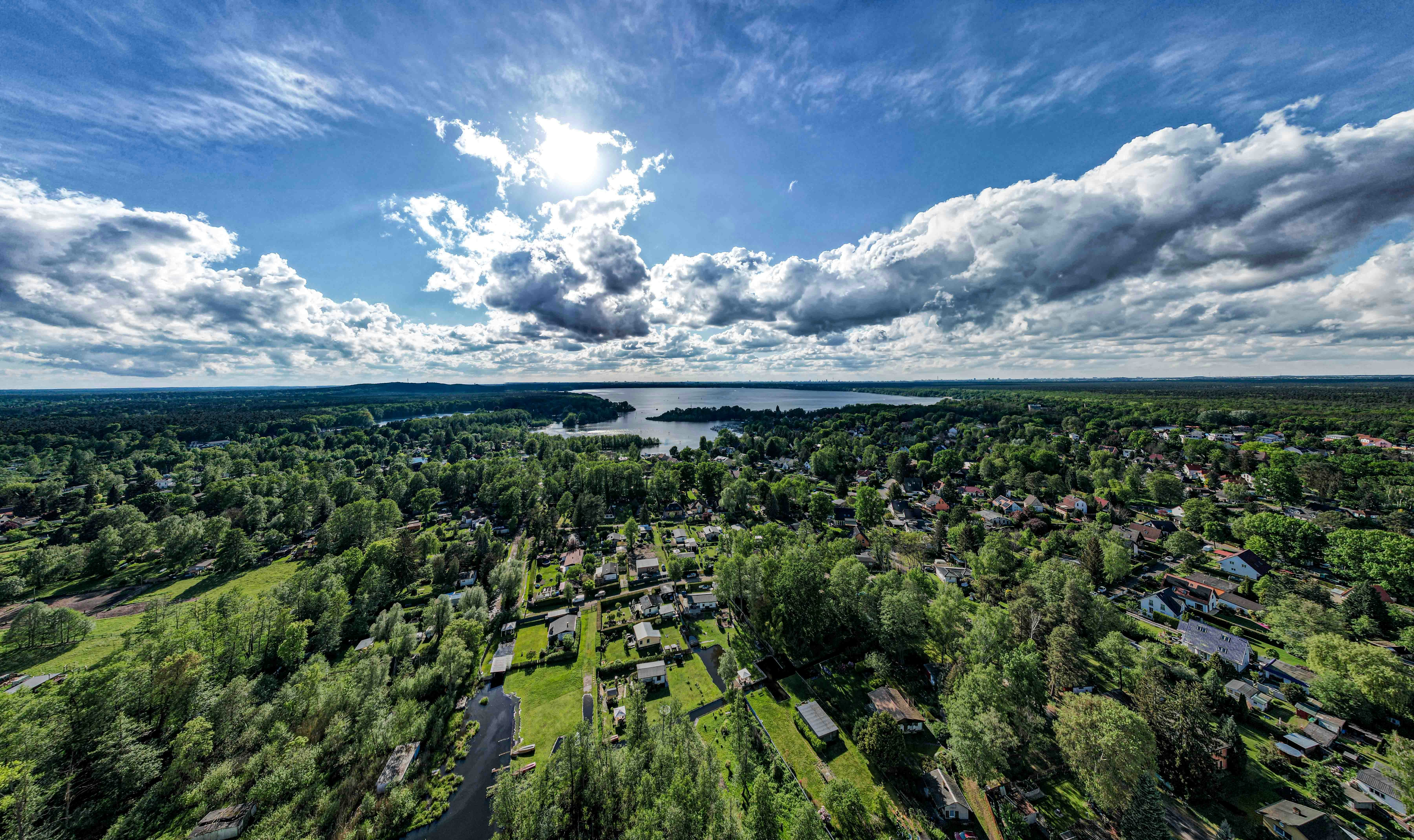 This expansive aerial shot captures a picturesque suburban landscape, where lush greenery envelops residential homes alongside a serene lake. The scene is set under a bright blue sky punctuated with dynamic clouds, casting dramatic shadows over the area. This idyllic setting highlights the harmonious blend of natural and urban elements, creating a tranquil environment that emphasizes community and natural beauty.