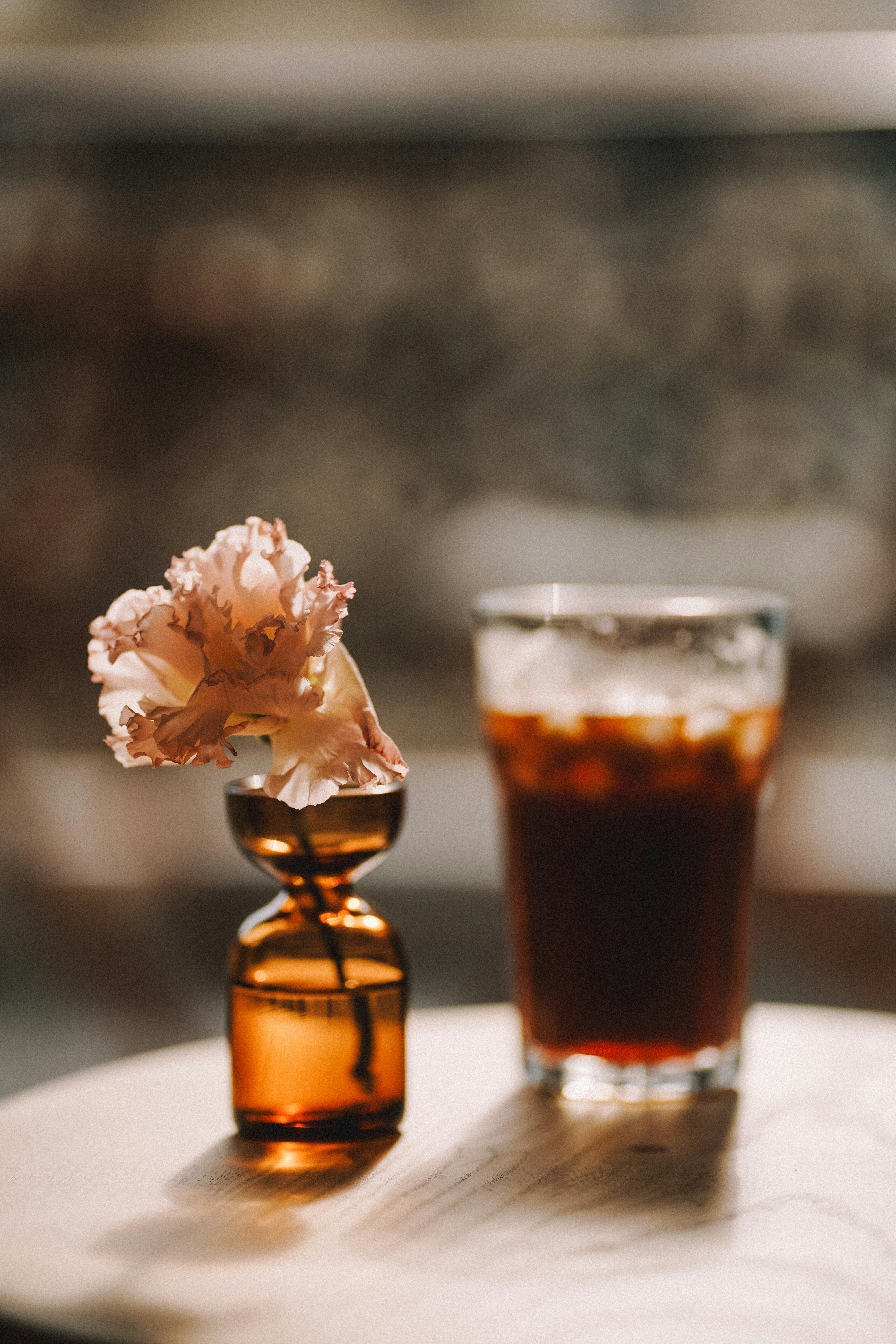 Small amber bottle with a pink carnation sits beside a tall glass of iced coffee on a marble surface, softly lit with a shallow depth of field.