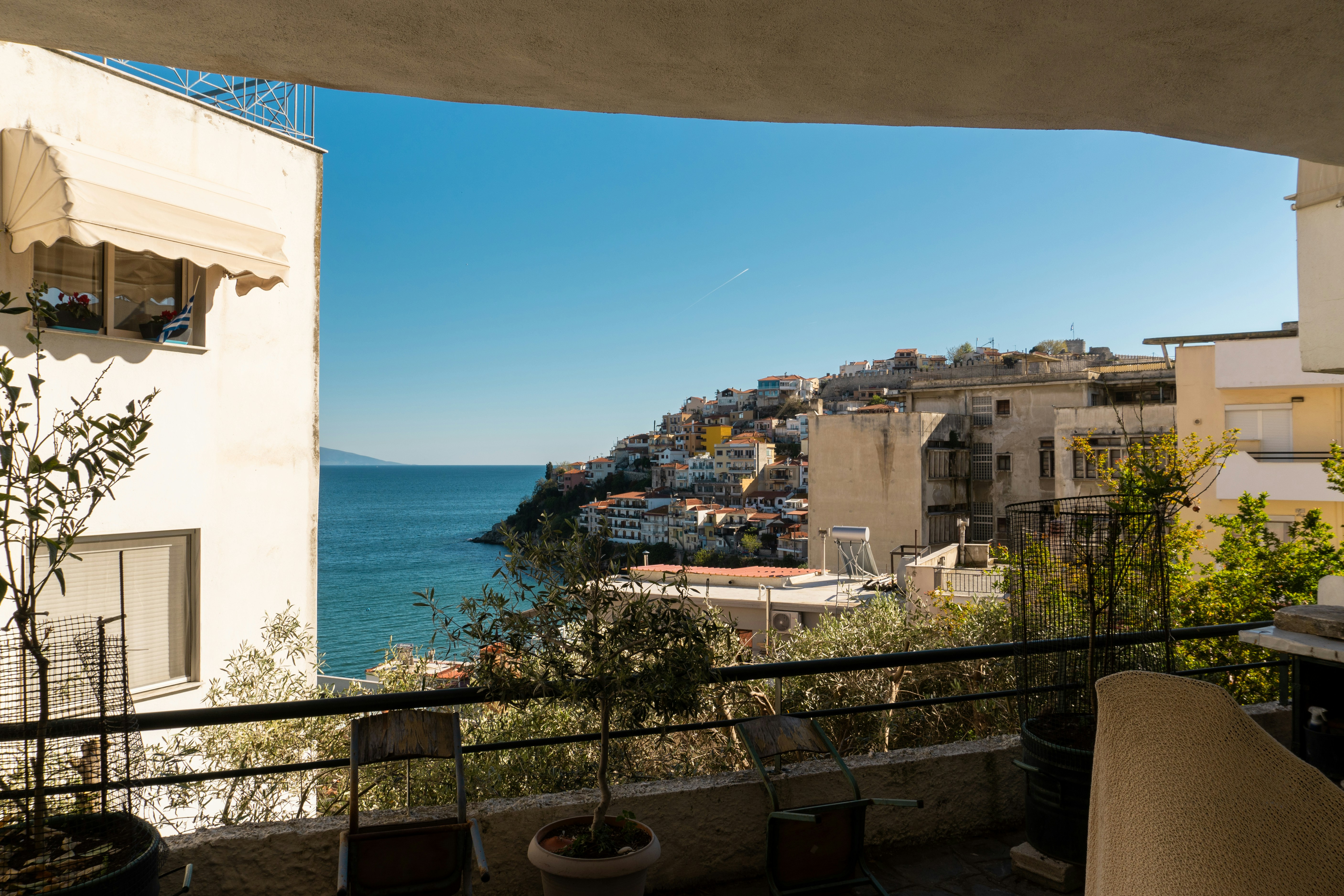 Sunlit balcony overlooking a serene coastal village with a clear blue sky and calm sea.