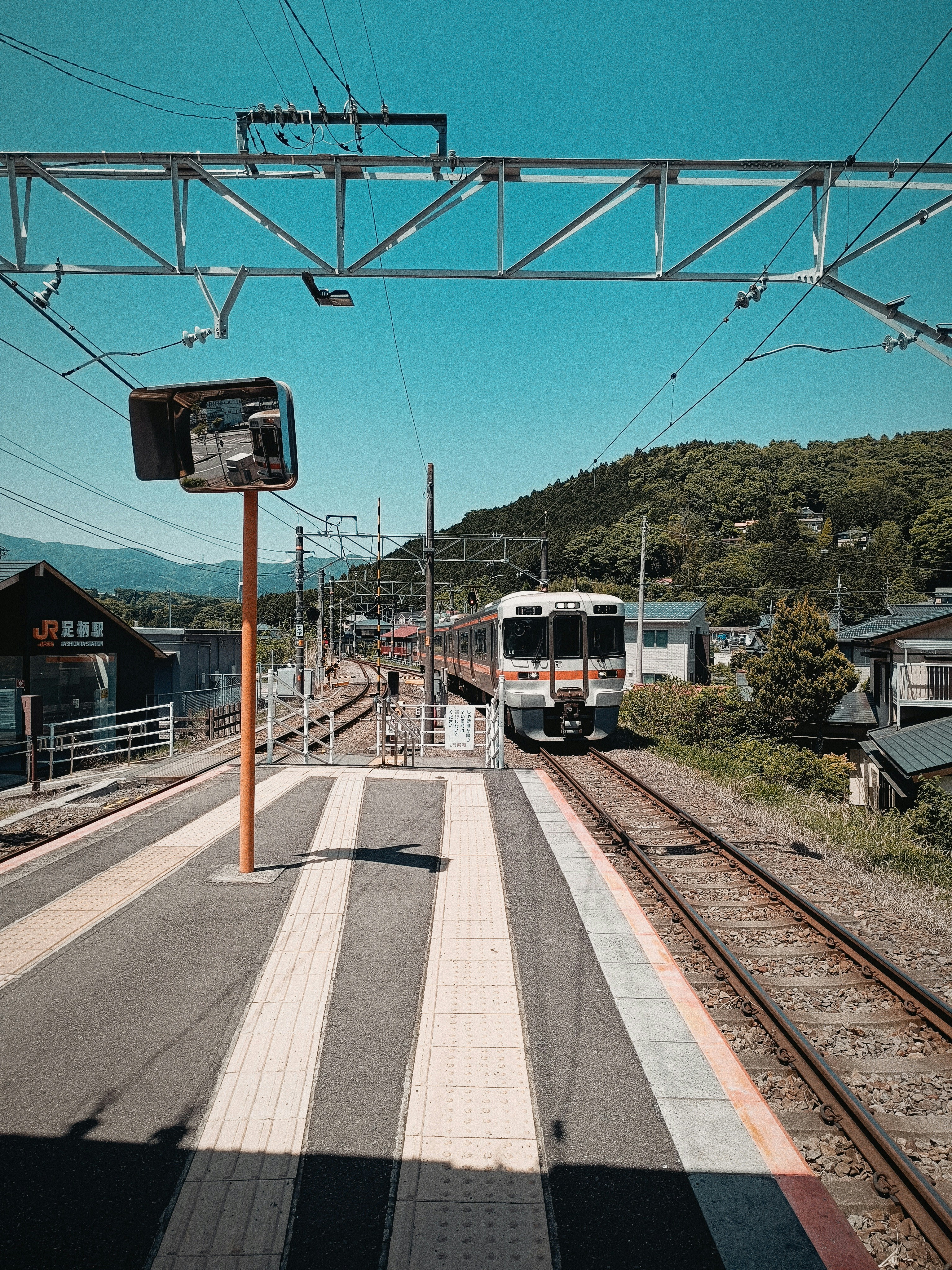 Sunlit rural platform with a commuter train approaching on electrified tracks, framed by overhead lines against a vibrant teal sky.