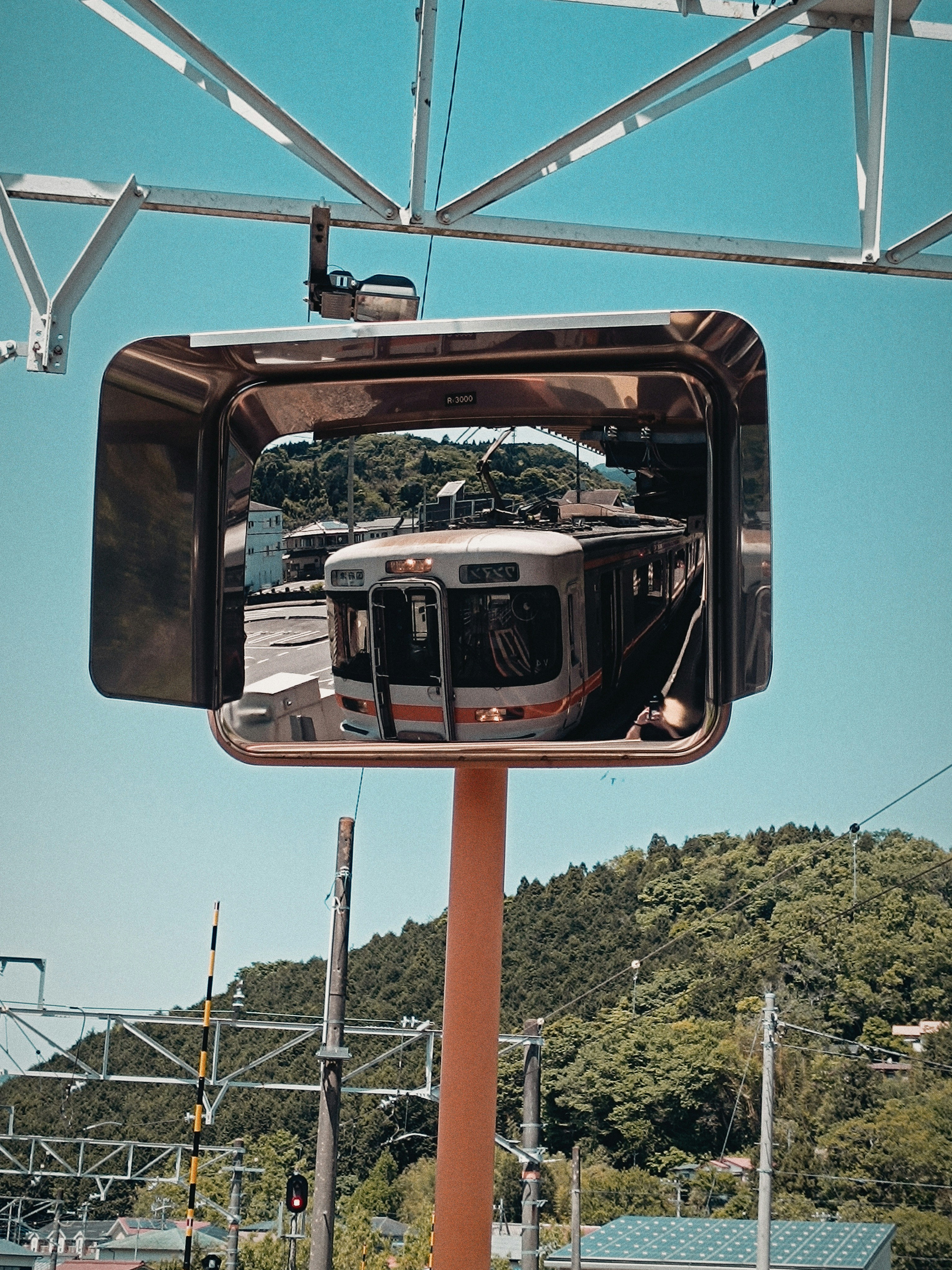 Front-on reflection of a train appears in a large rectangular mirror mounted above the tracks, framed by blue sky and structural supports.