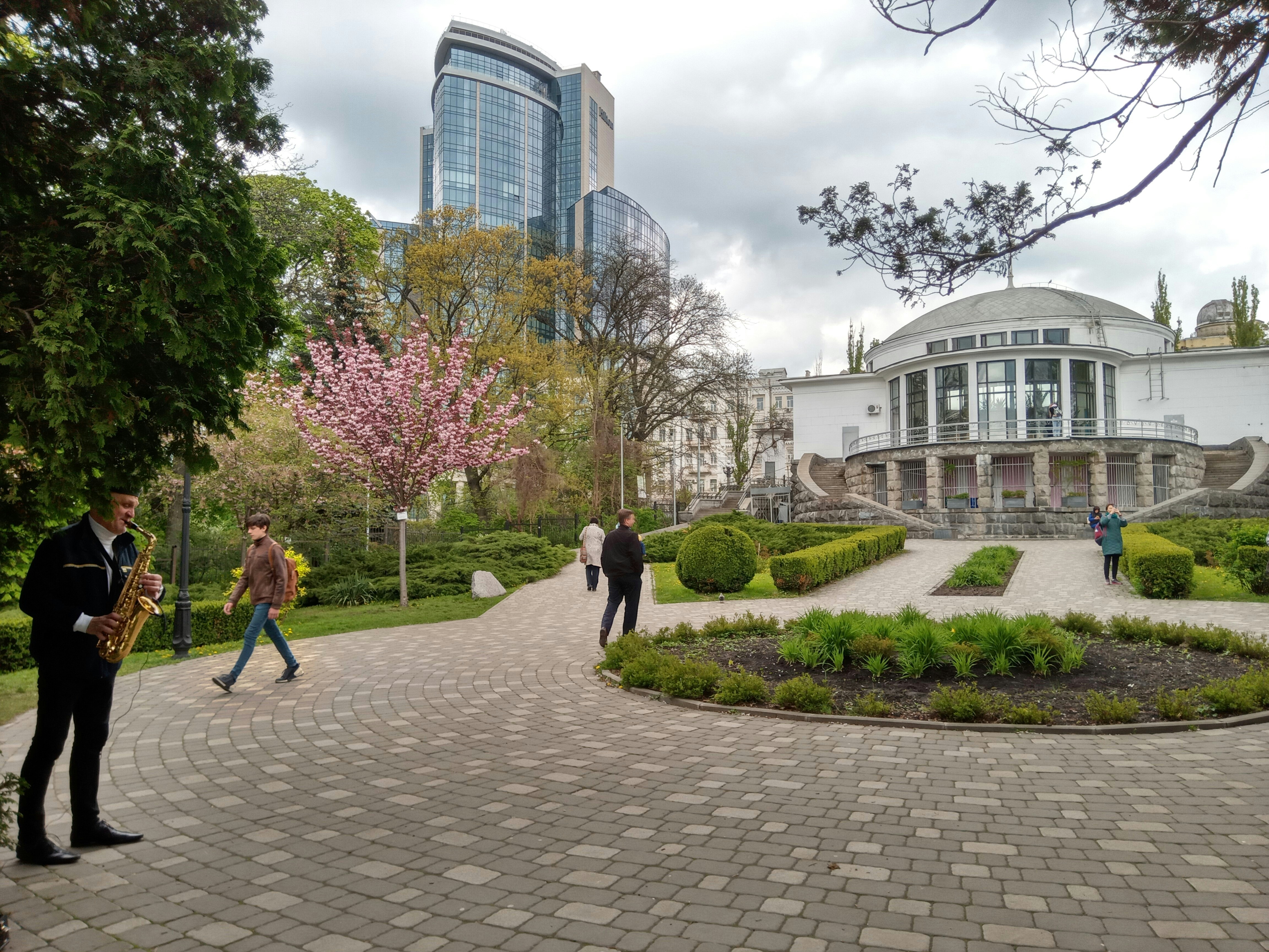 Musician playing saxophone in a vibrant park surrounded by blooming cherry trees and modern architecture. People stroll along the winding path.