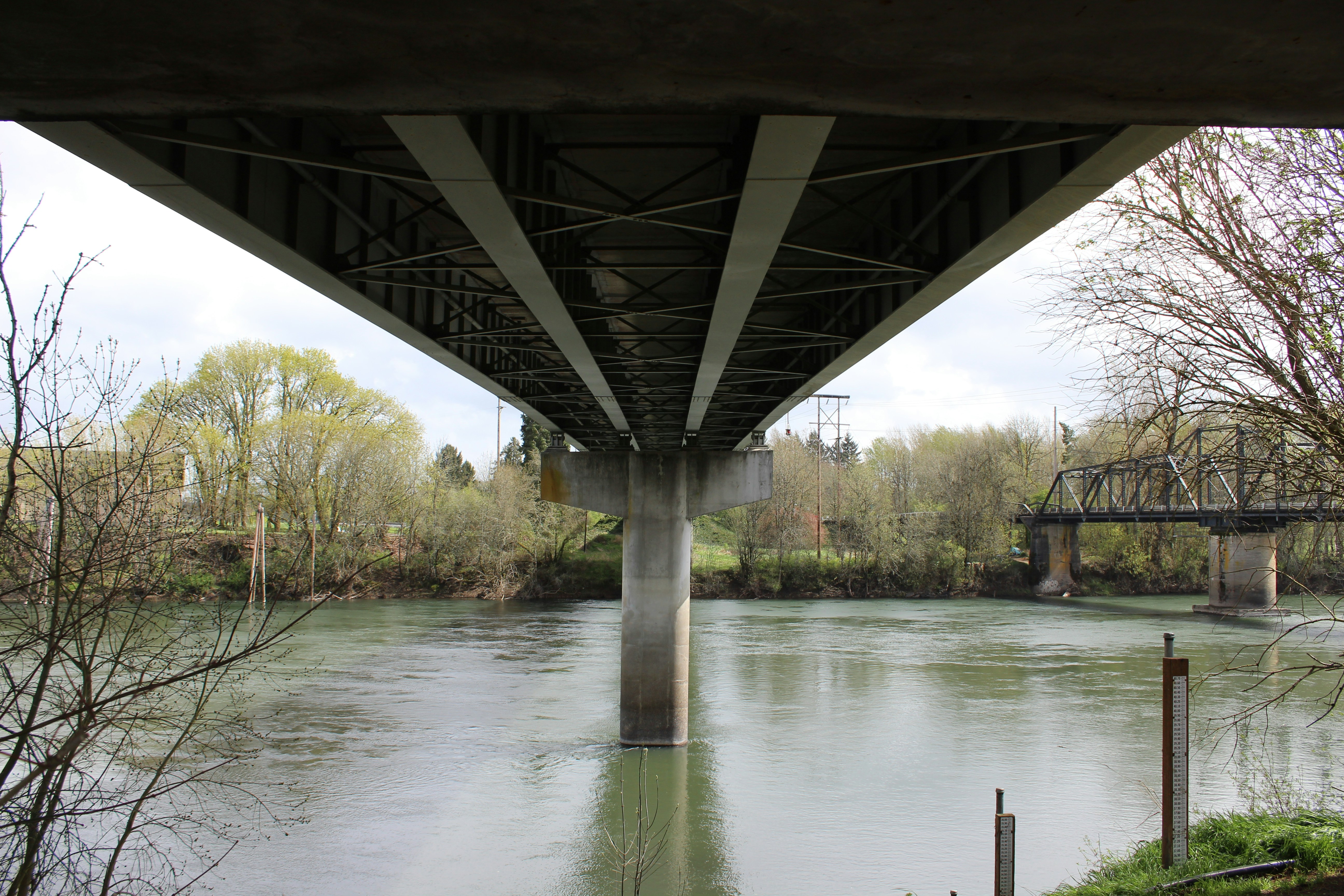 The view from beneath a bridge, showcasing the intricate steel structure overhead and the calm river flowing below.