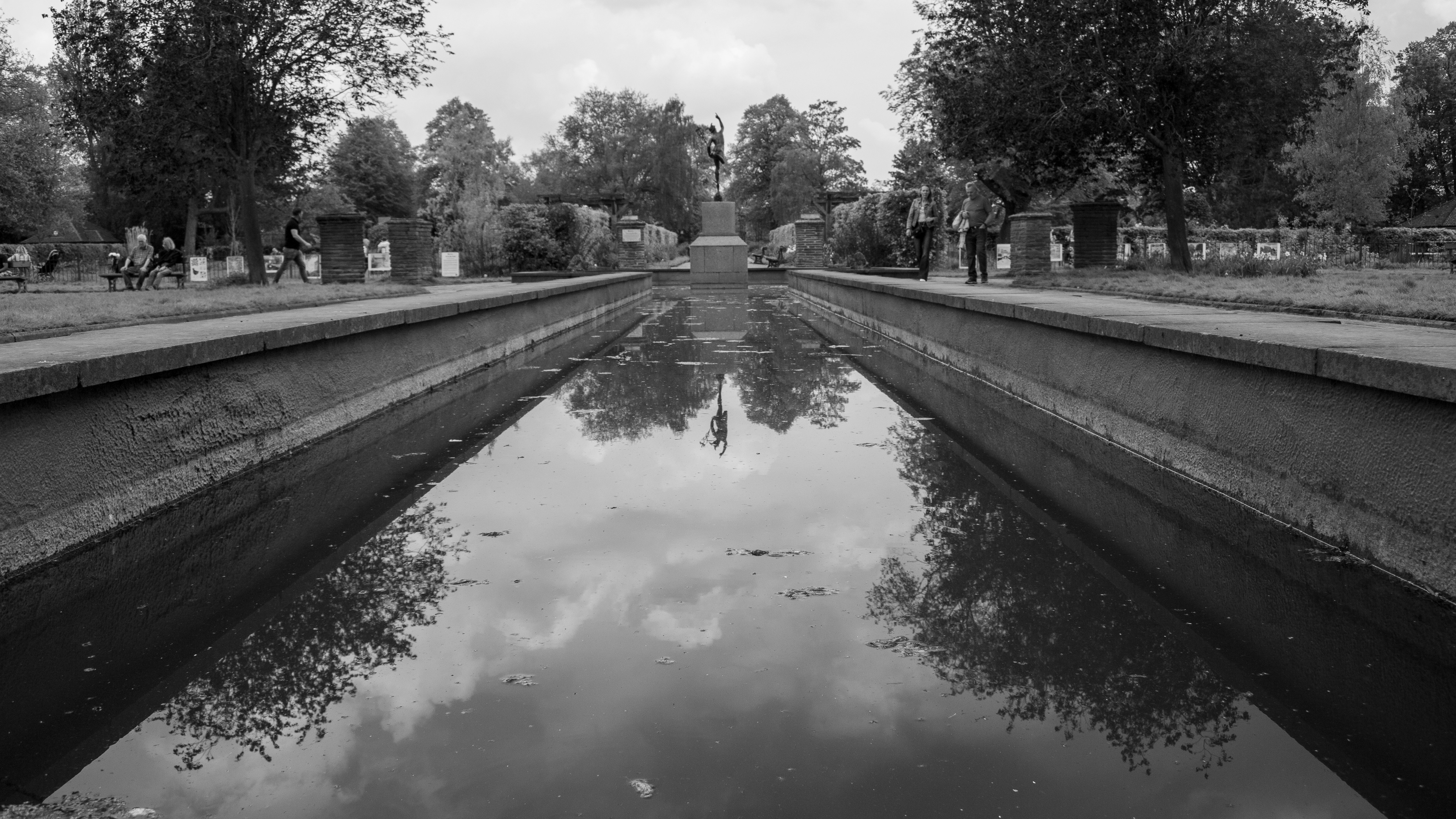A tranquil park scene featuring a central statue reflected in a calm waterway, framed by lush trees and pathways. The monochrome tones enhance the peaceful atmosphere.