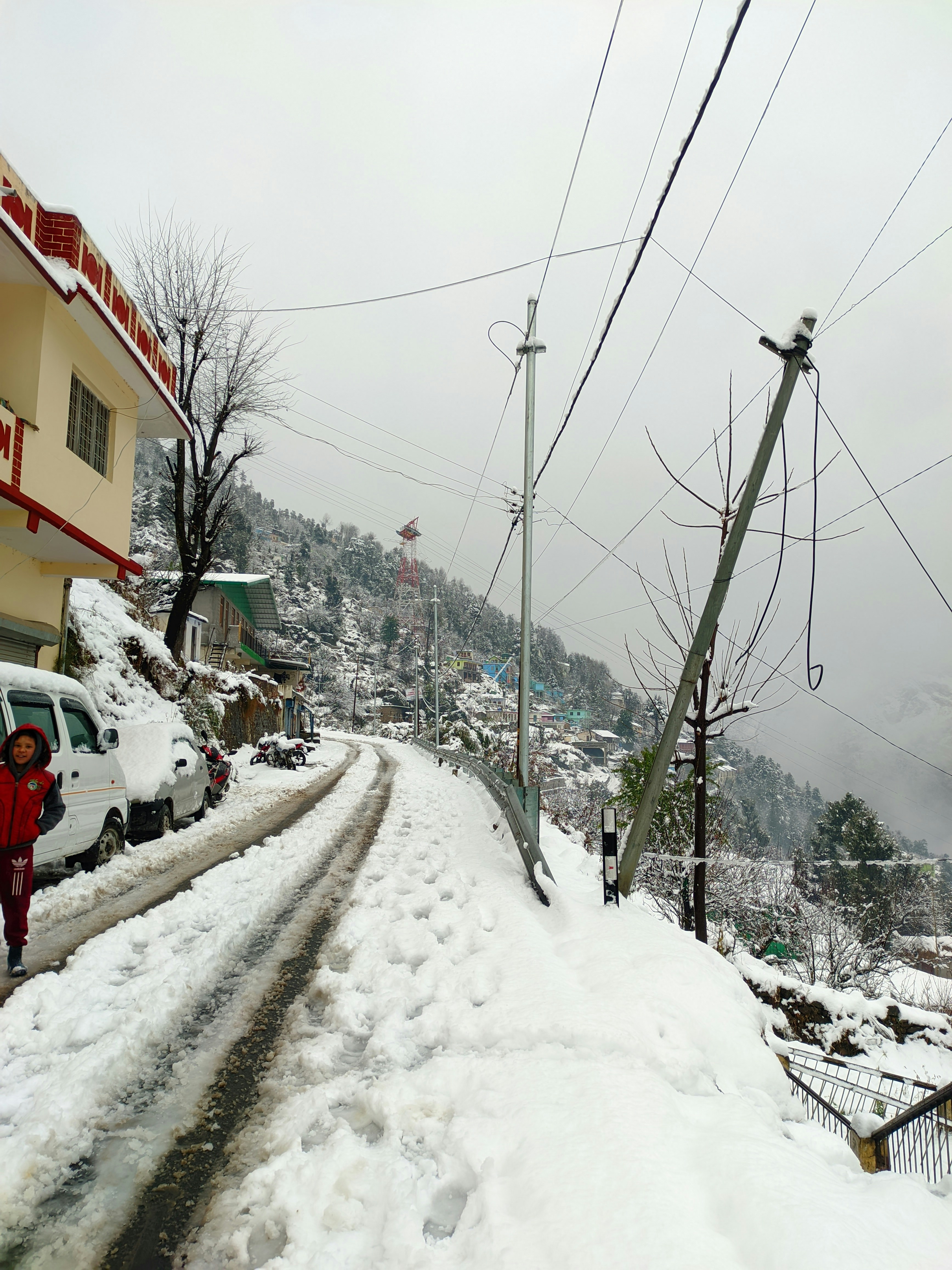 Snow-covered hillside street lined with houses and leaning power poles, with a person in a red jacket near the left curb. A distant village rises into the gray sky.