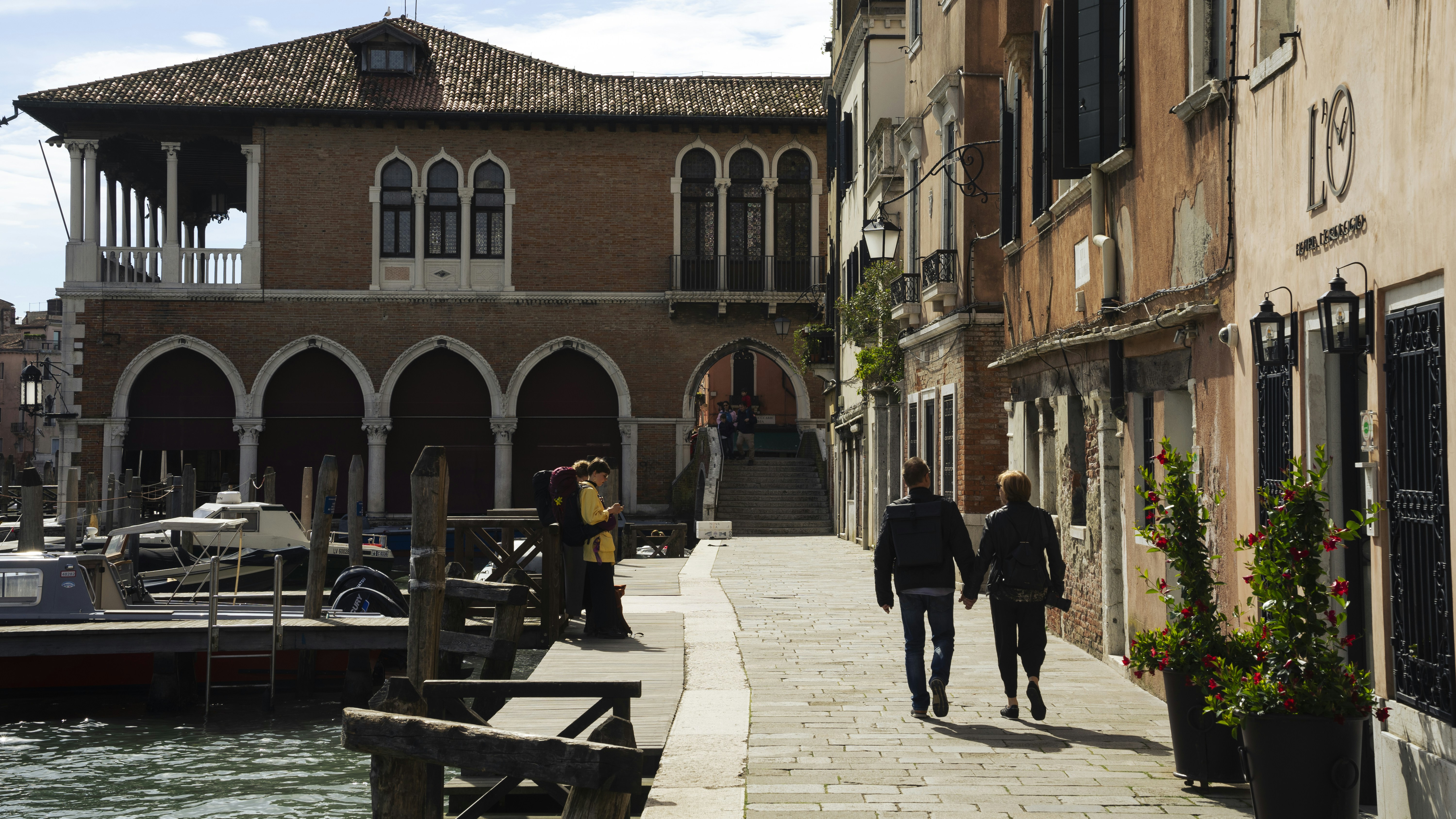 Two people walk along a sunlit canal in Venice, bordered by historic buildings and moored boats.