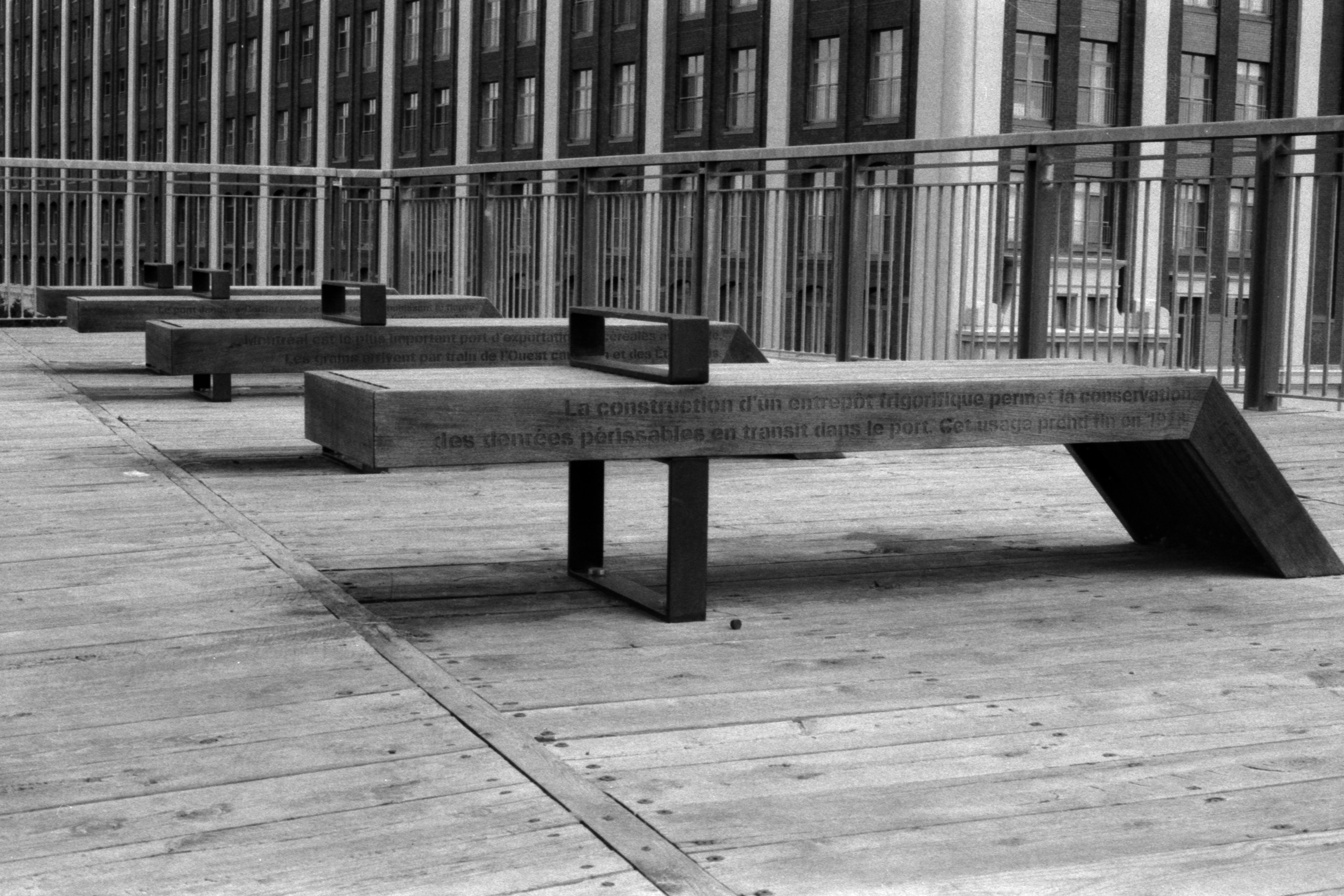 A black-and-white photograph of an urban plaza featuring angular benches, metal rails, and expansive concrete planes. The composition emphasizes minimalist geometry and empty space.