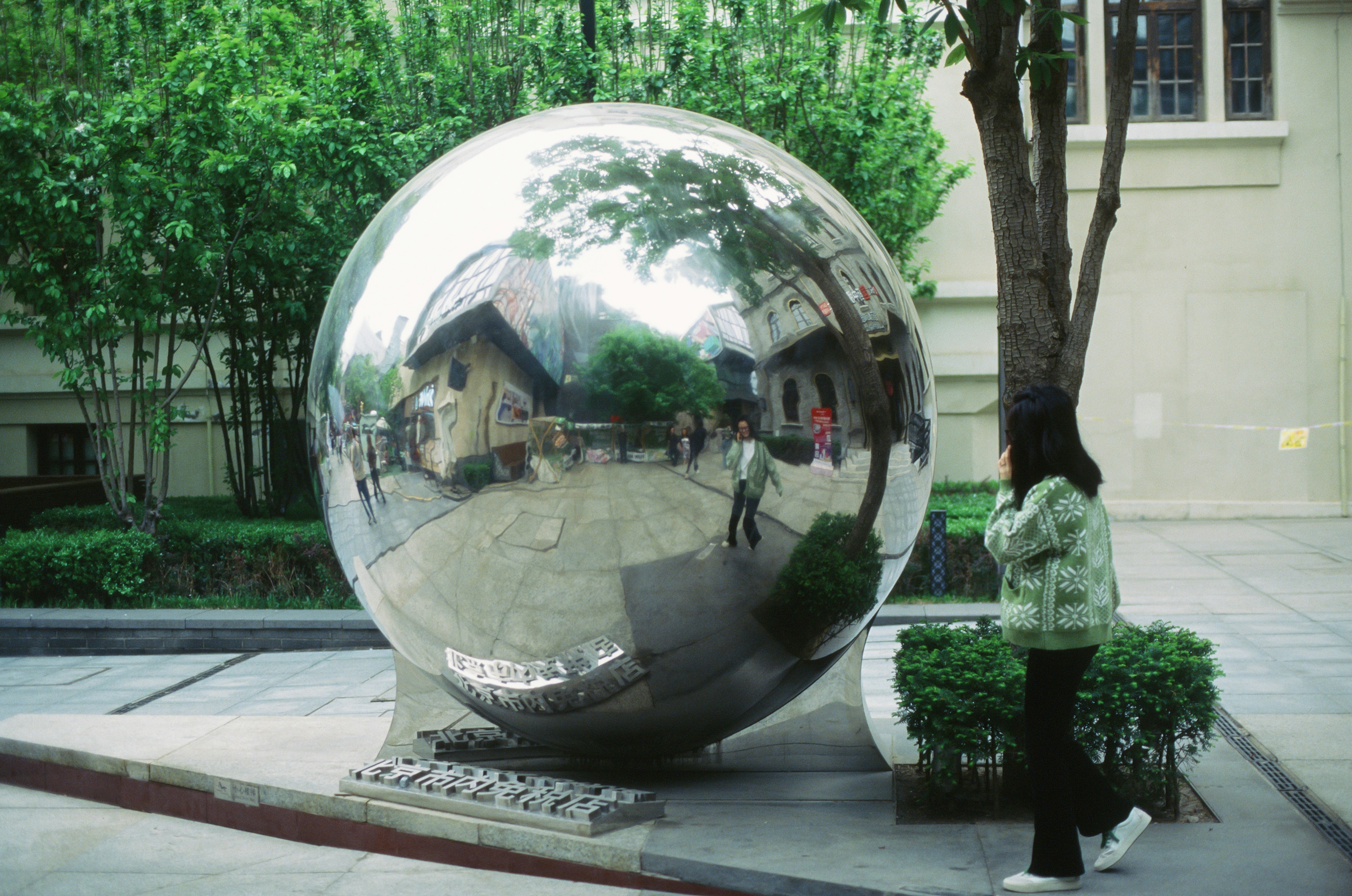 Reflective sphere in a courtyard mirrors buildings and passersby amidst lush greenery.