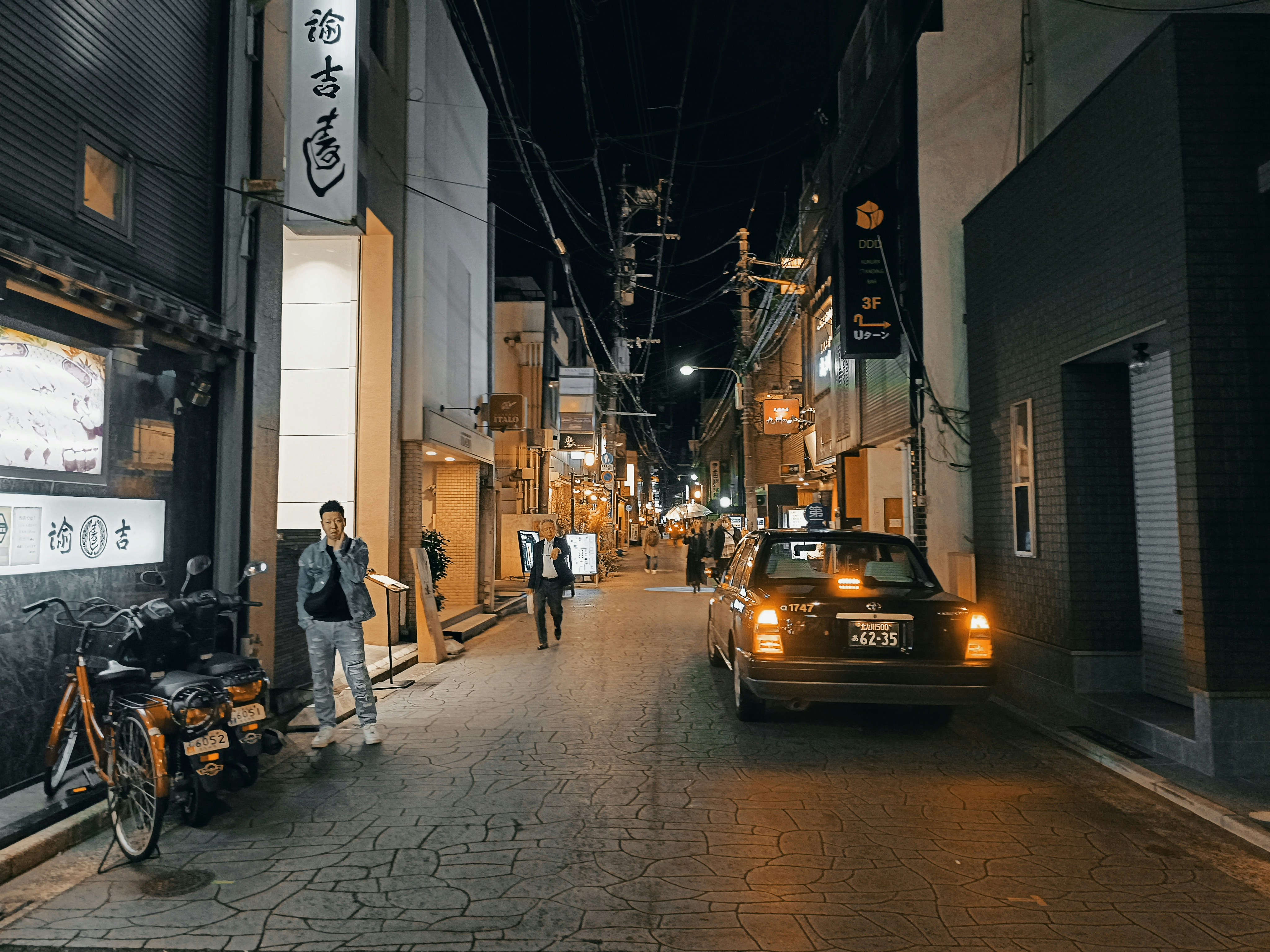 Nighttime urban street scene with a car parked beside a narrow building and a few pedestrians walking.