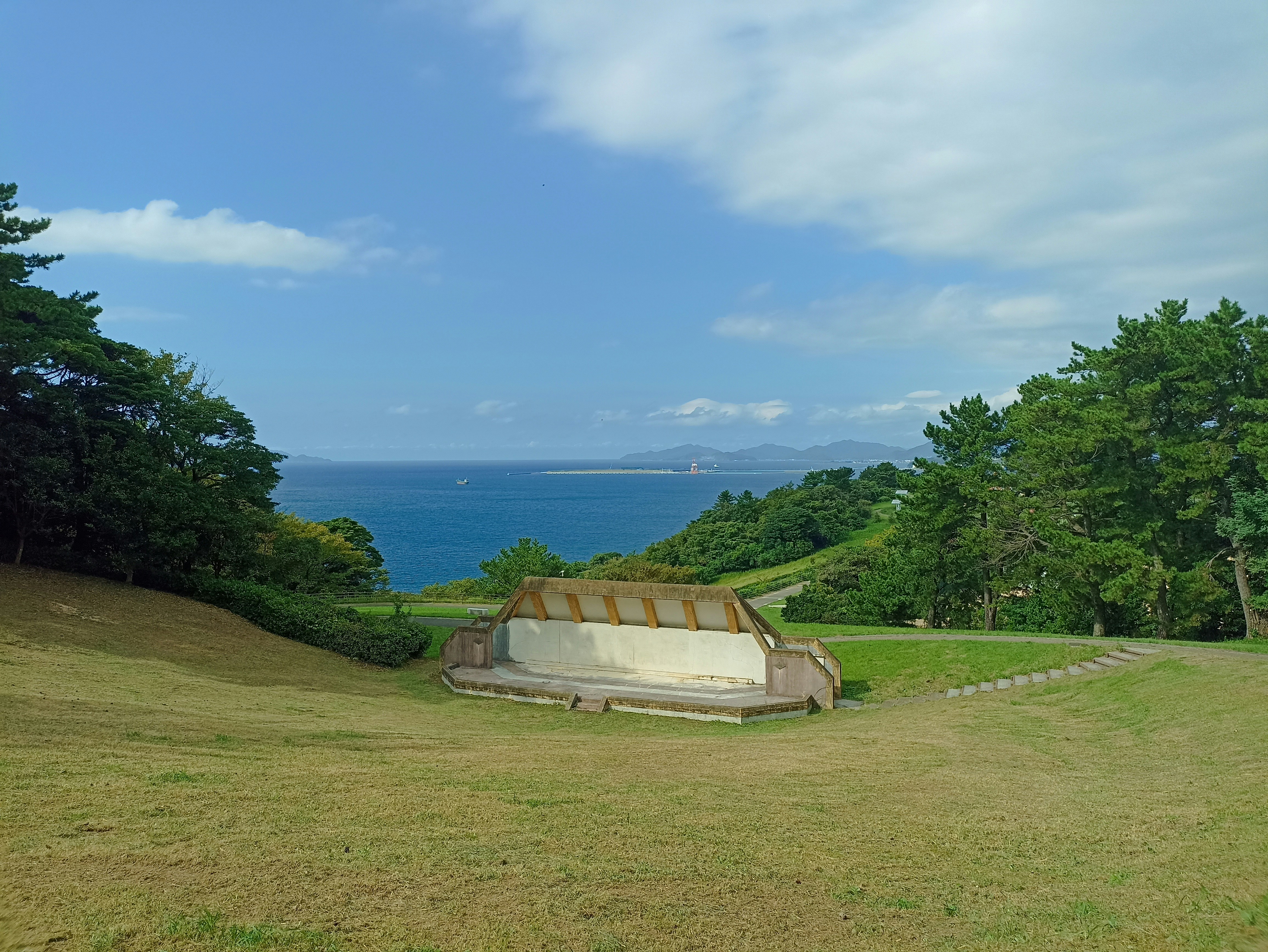 Wooden bench atop a grassy hill overlooking a tranquil blue sea under a partly cloudy sky.