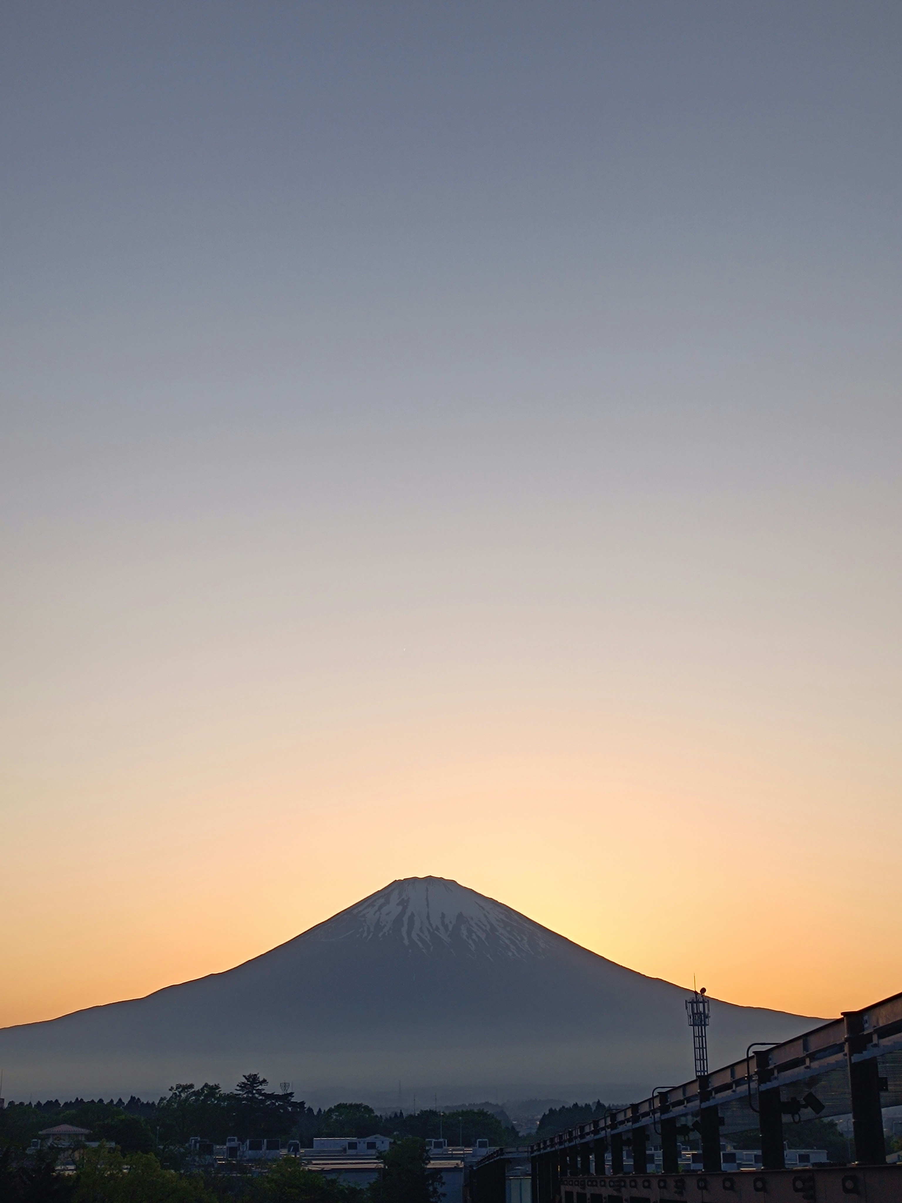 Snow-capped volcanic summit rises above a low-lying town as dawn paints the sky in warm gradients. In the foreground, a raised walkway and railing guide the eye toward the tranquil peak.