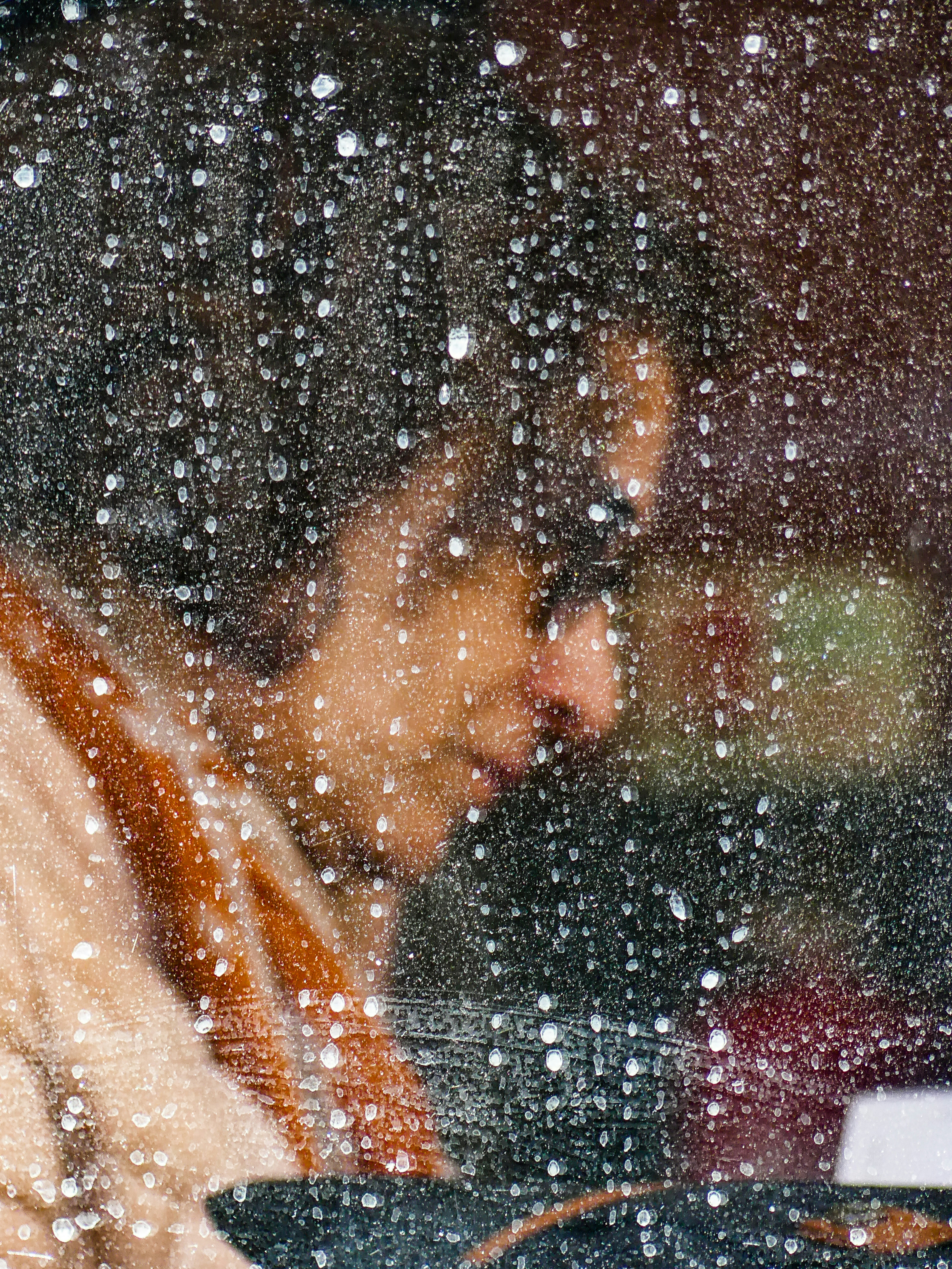 Person viewed through rain-speckled glass focusing on a laptop.