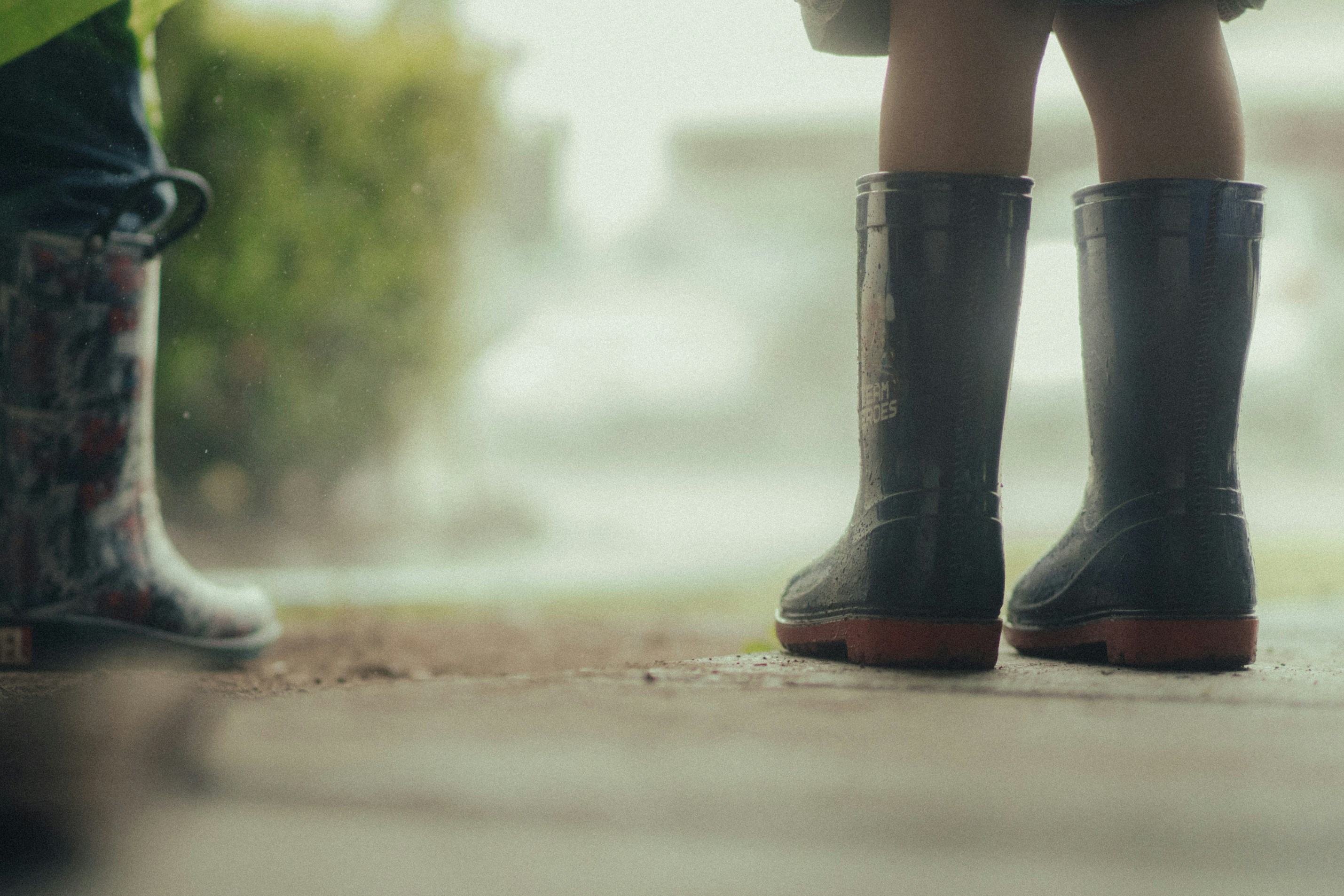 Children wearing rubber boots standing on a wet sidewalk with blurred greenery in the background.
