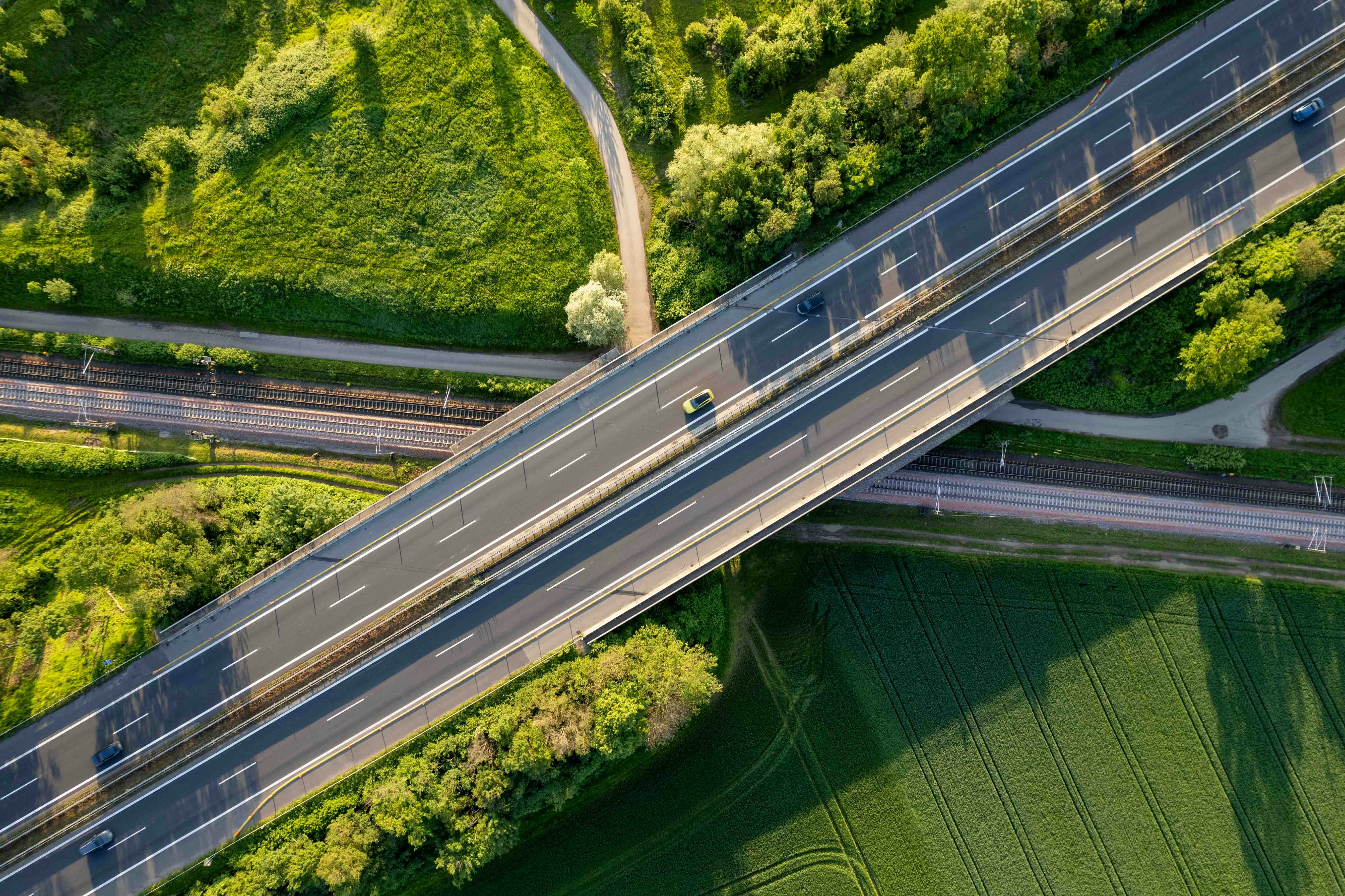 This aerial image captures a dynamic scene where a busy highway crosses over railway tracks, surrounded by lush green agricultural fields. Vehicles move along the highway, showcasing everyday mobility and connectivity. The contrast between the engineered straight lines of the road and railway against the natural patterns of the green fields highlights the intersection of human activity and natural landscapes.