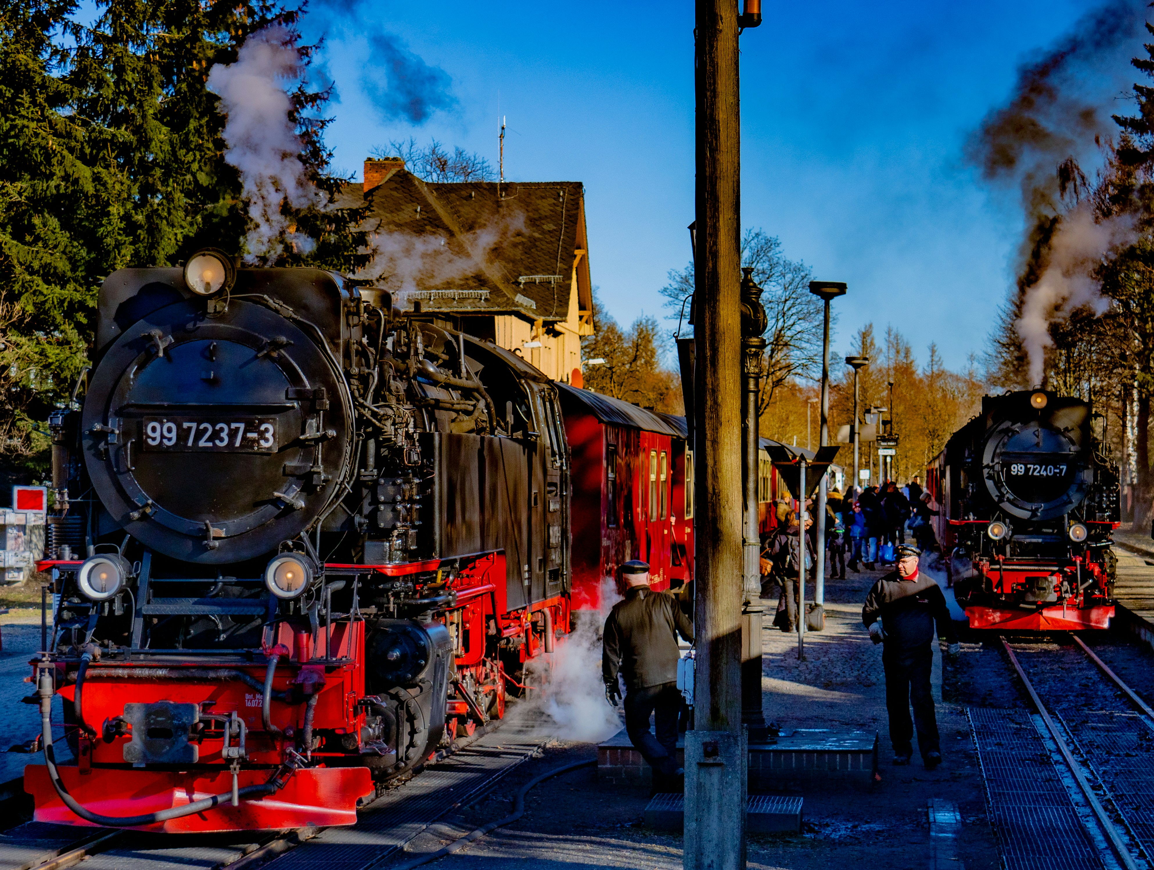 Two steam locomotives emit plumes of smoke at a vibrant train station under a clear blue sky.